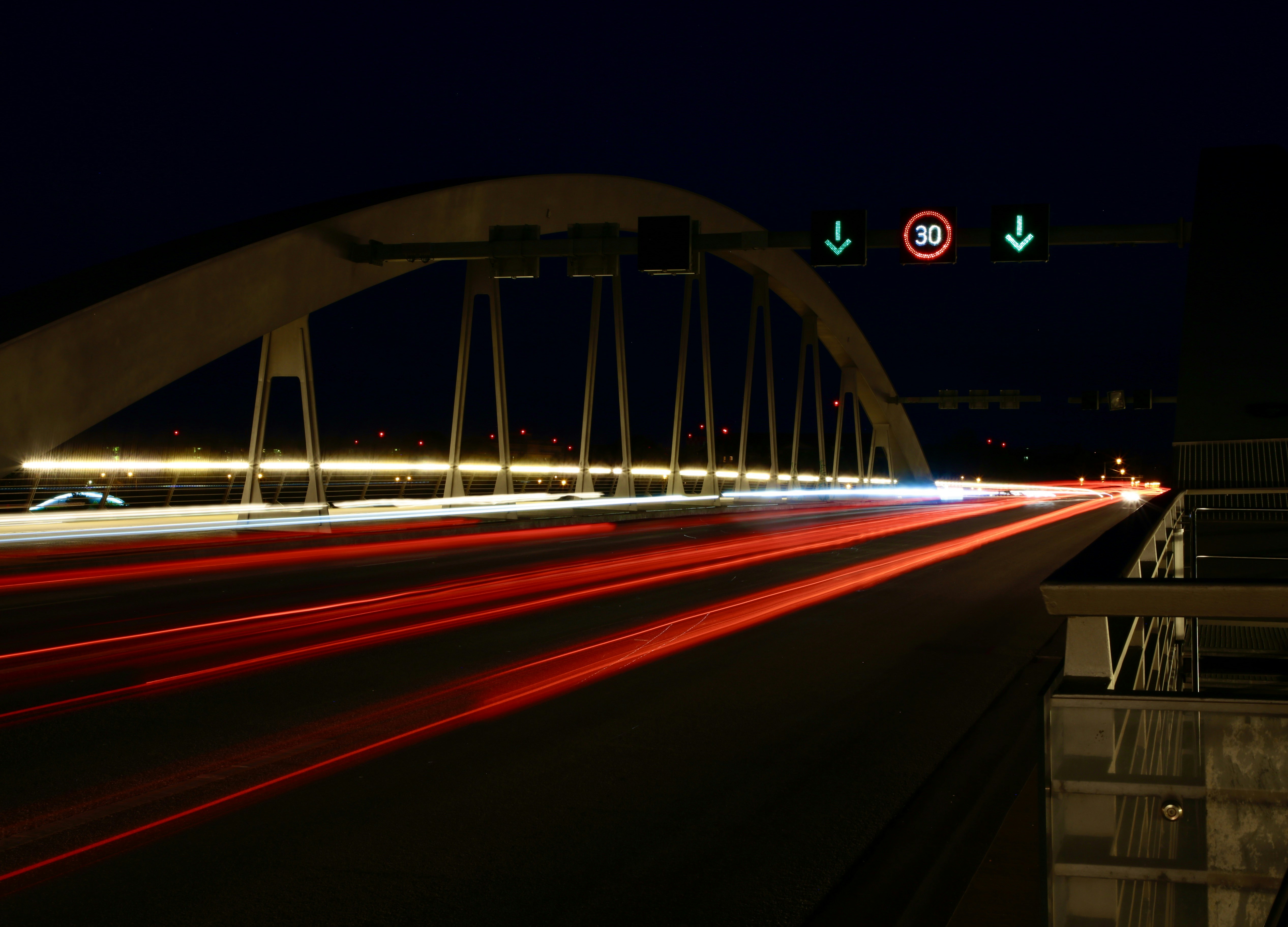 Light trails from vehicles crossing an illuminated bridge at night, with traffic signs visible above. The scene captures the essence of urban movement and structure.