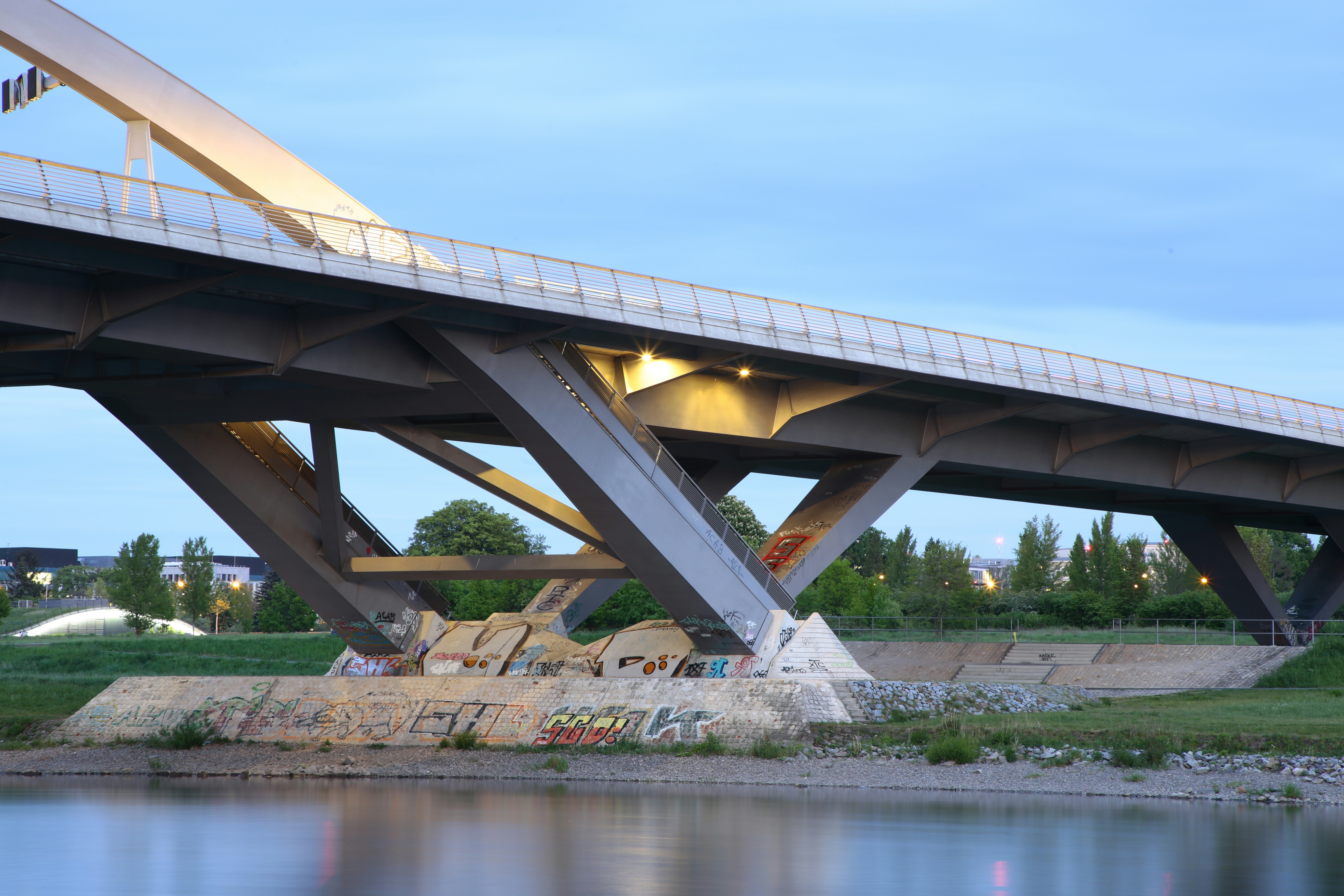 Modern bridge structure supported by geometric pillars, adorned with graffiti along the riverbank, reflecting urban art and nature's tranquility.