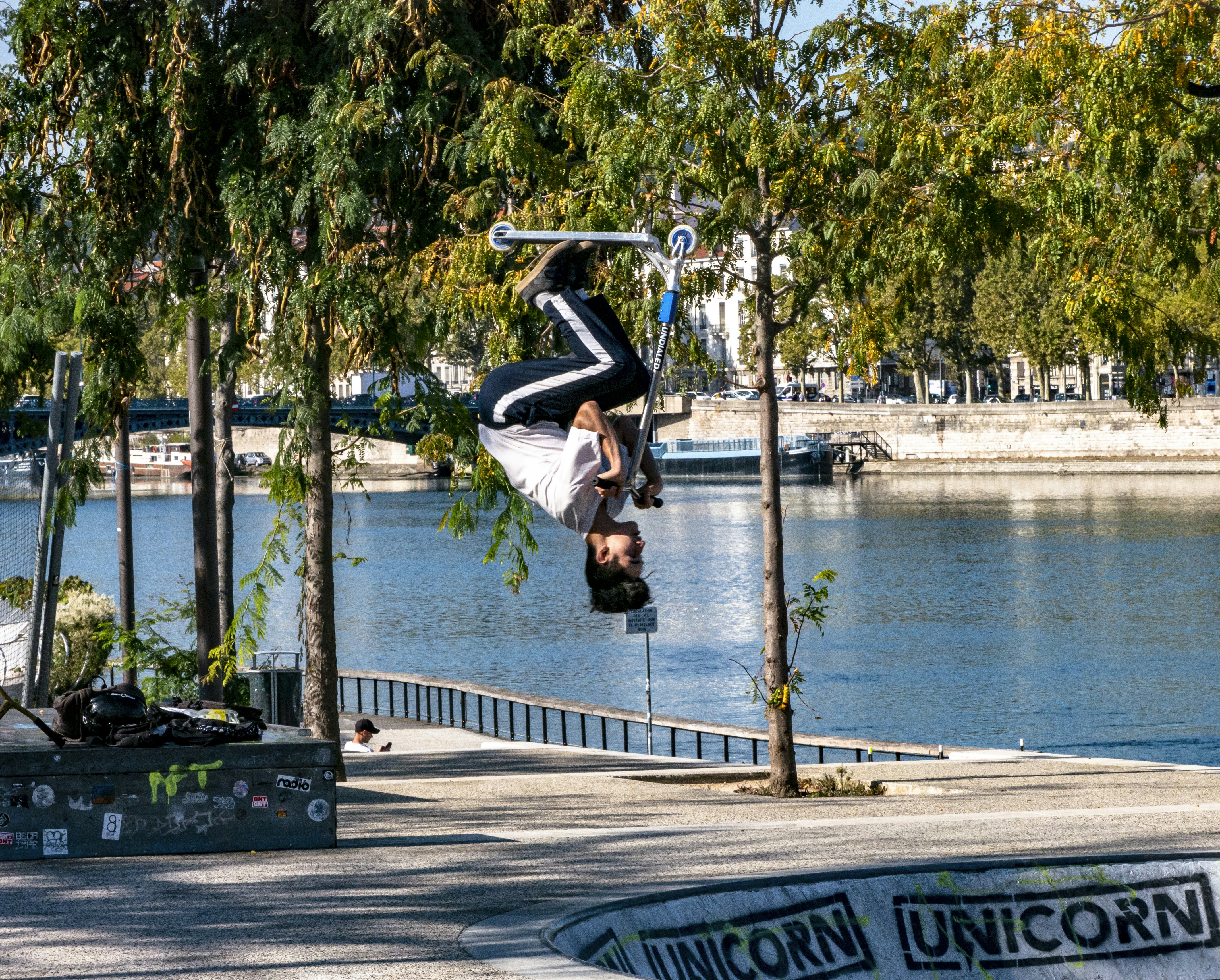 Scooter rider performing an inverted aerial trick in a skatepark near a calm river, surrounded by trees.