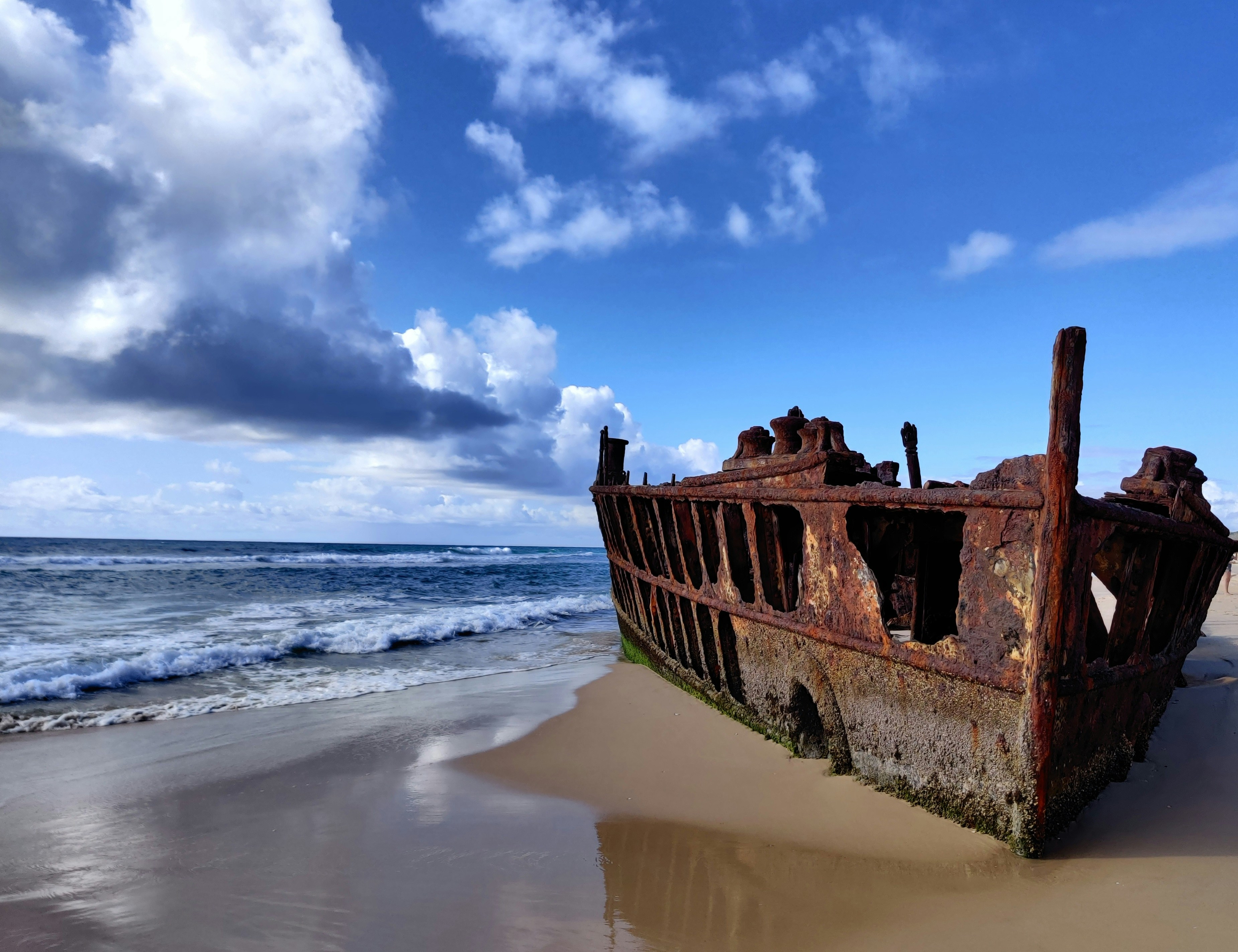 Weathered shipwreck resting on sandy beach, with waves gently lapping at its hull under a bright blue sky.