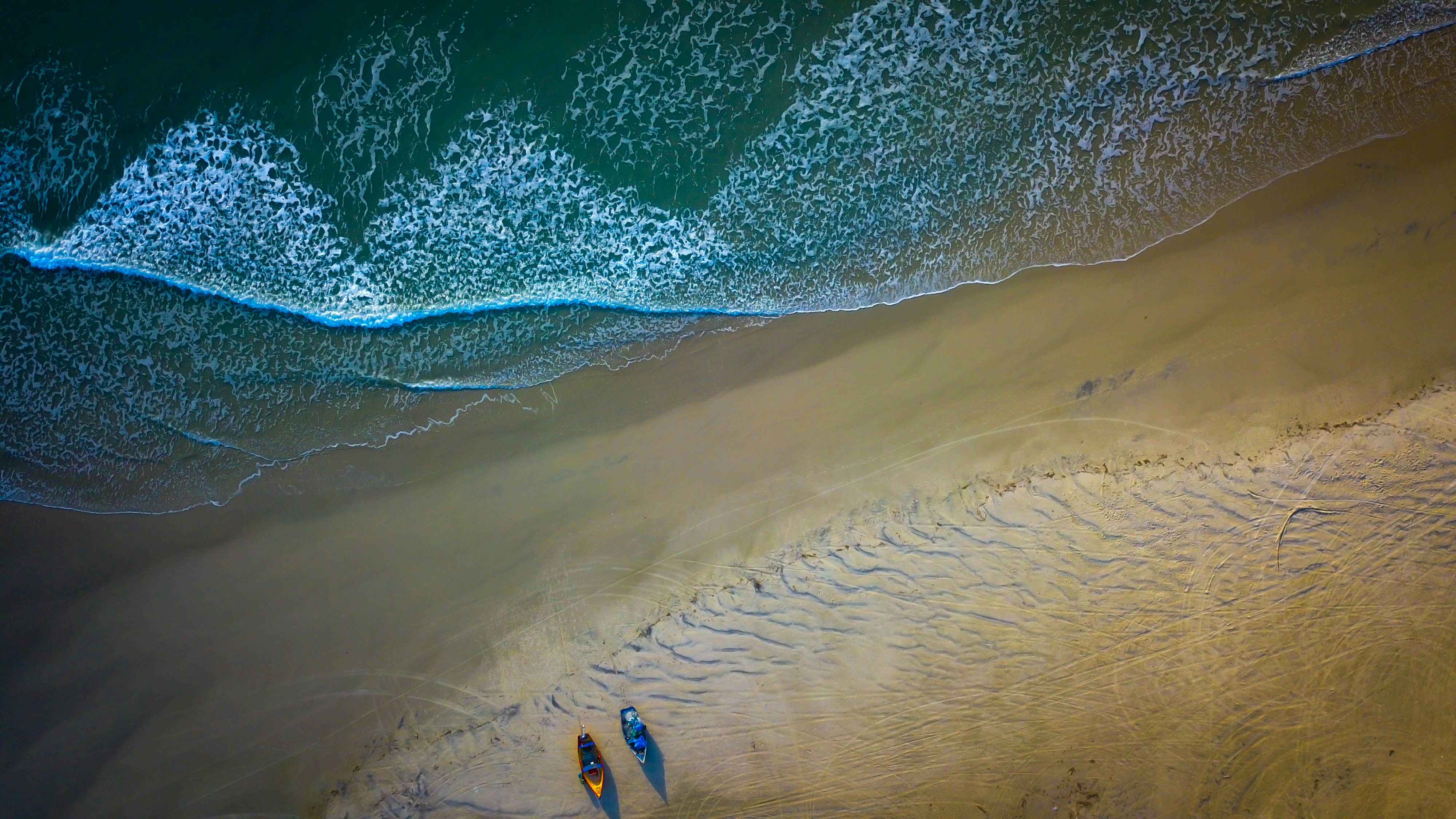 Aerial view of ocean waves gently lapping a sandy beach, with two kayaks on the shore.