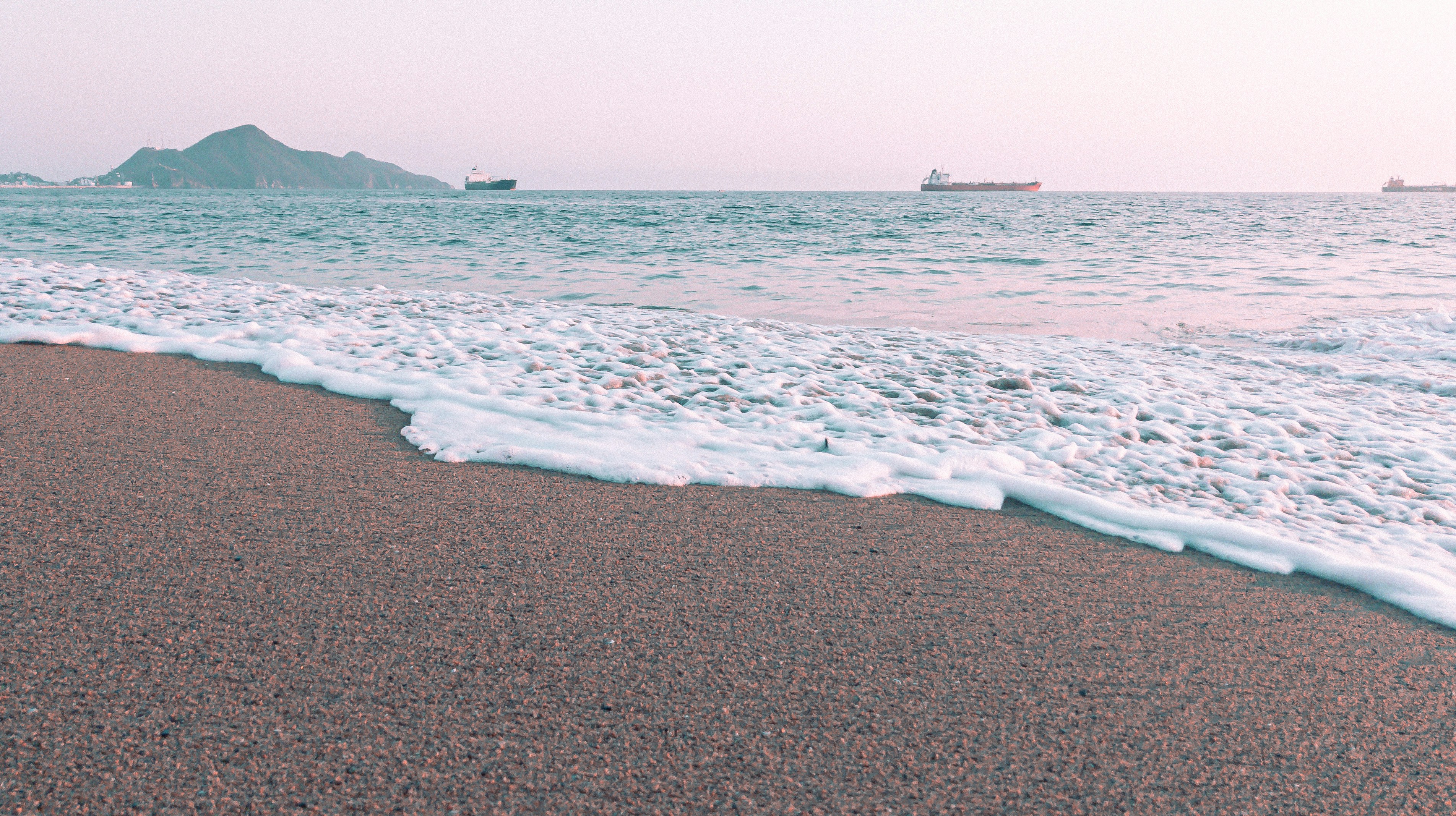 Gentle waves caress a sandy beach with distant ships on the horizon at dusk.