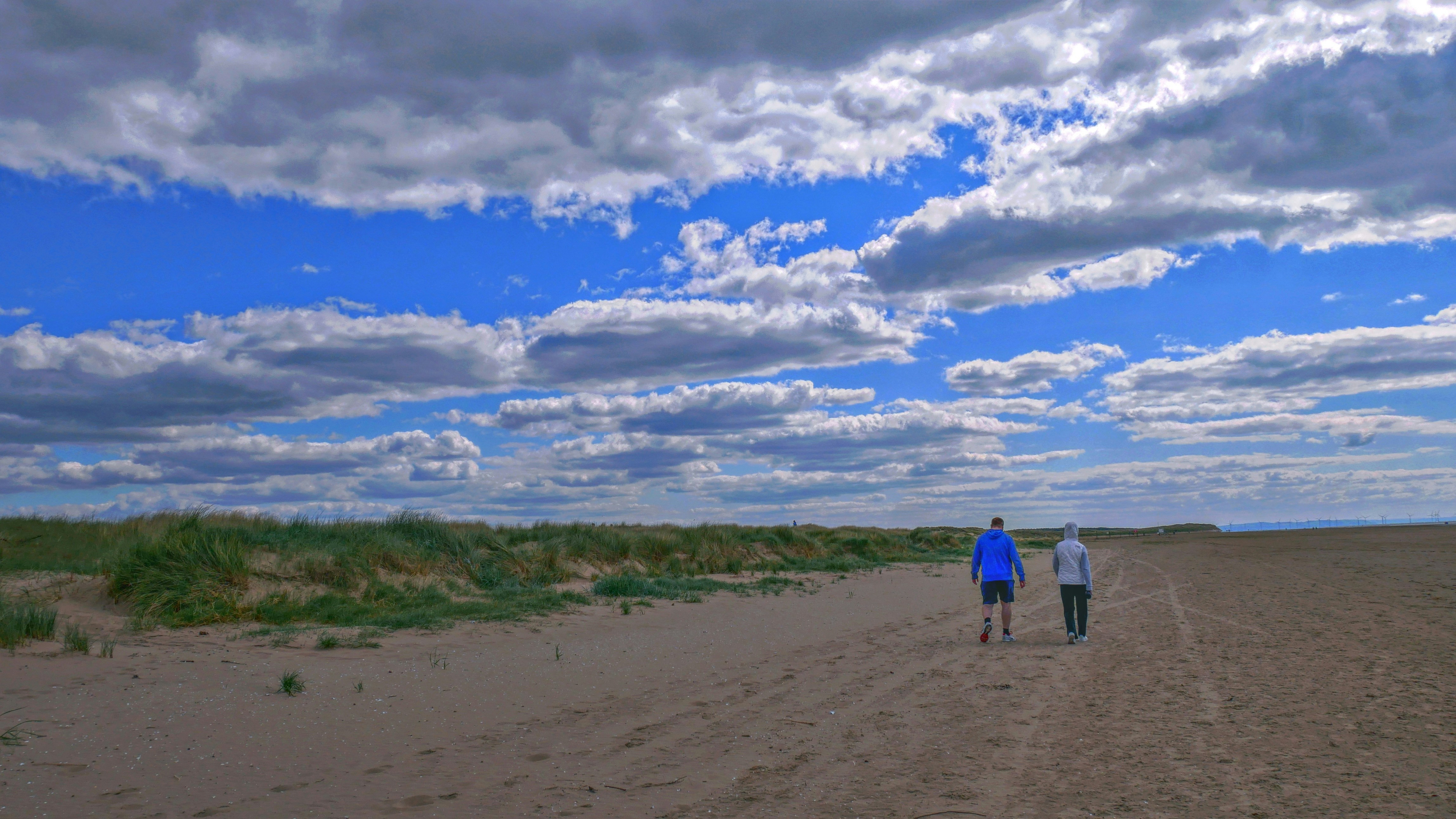 Two figures stroll along a sandy beach under a vast sky filled with dynamic clouds. The contrast between the earth and sky creates a sense of tranquility.