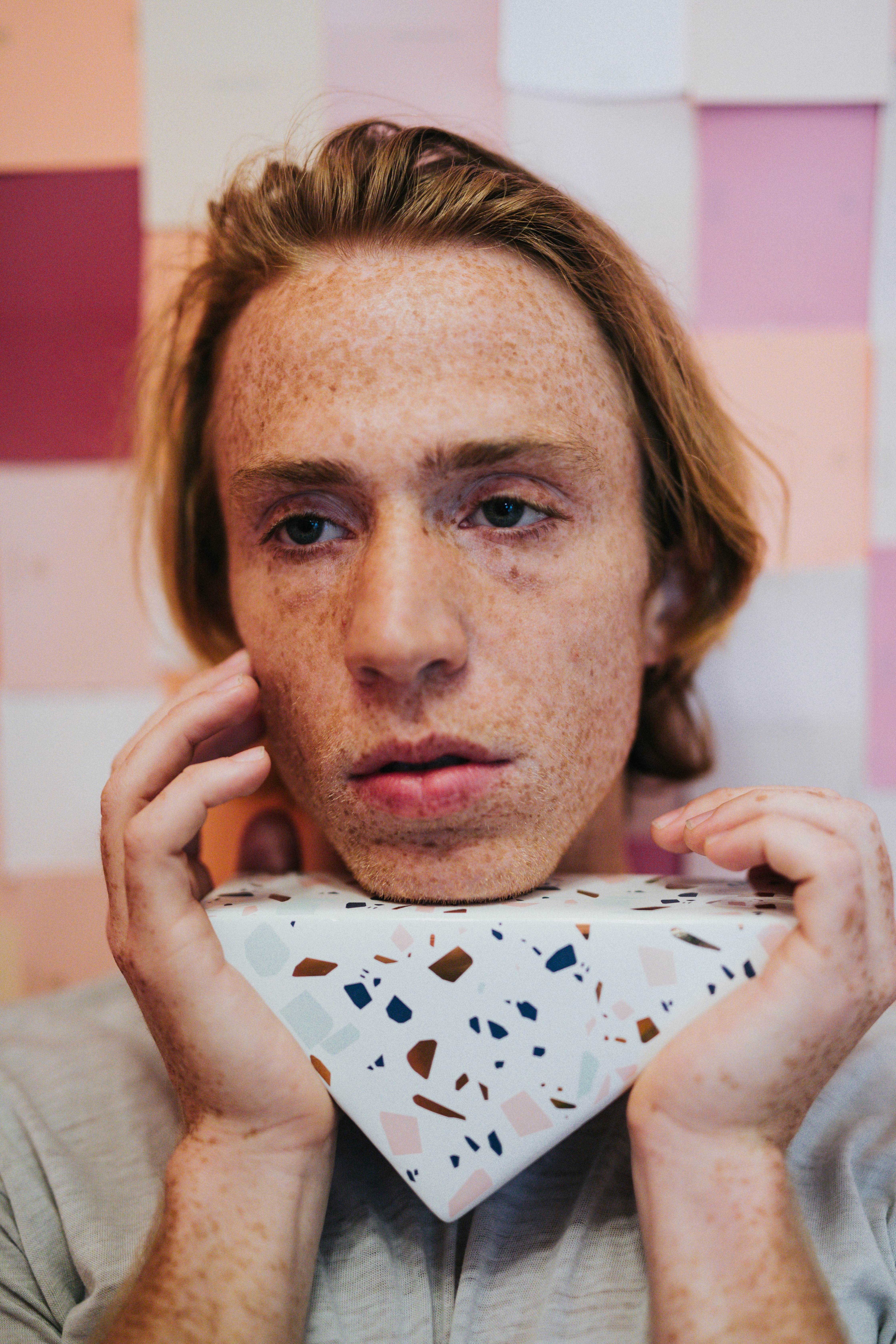 A young man with freckles holds a geometric object close to his face, set against a colorful patchwork background of pastel hues.