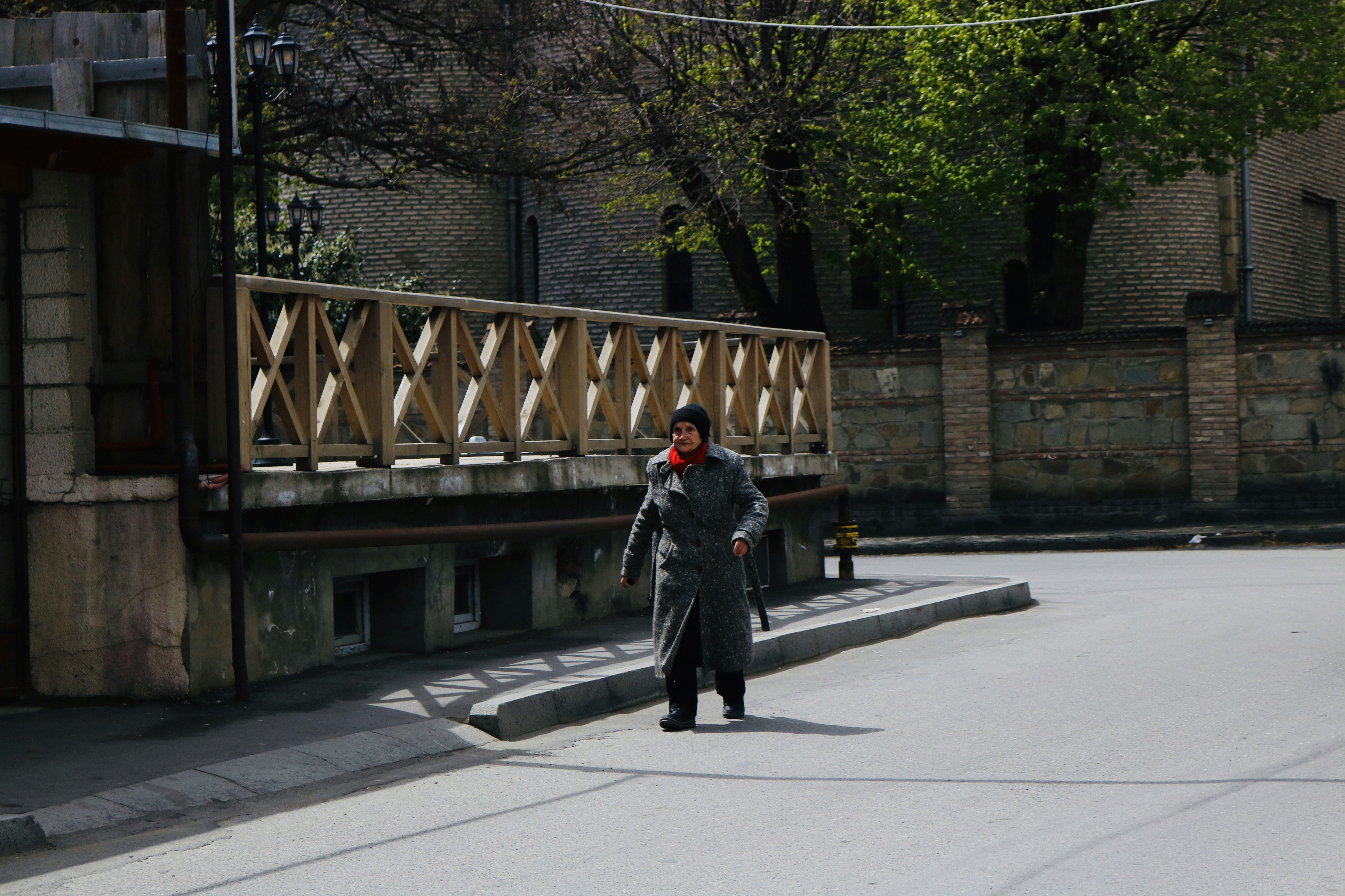 An elderly woman walks along a quiet street, dressed in a long coat with a vibrant scarf, amidst a backdrop of urban architecture and greenery.