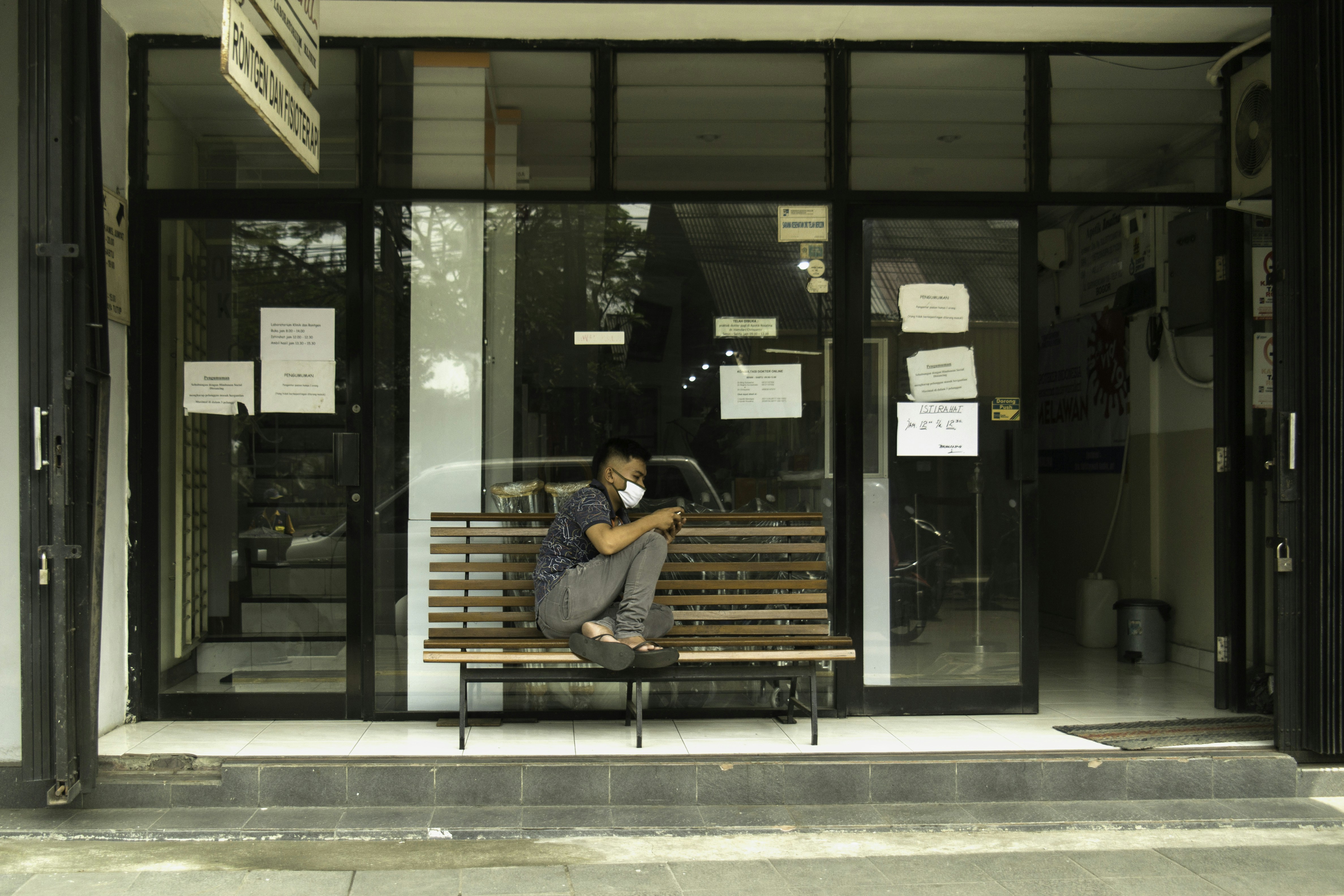 Person seated on a bench using a cellphone outside a closed pharmacy.