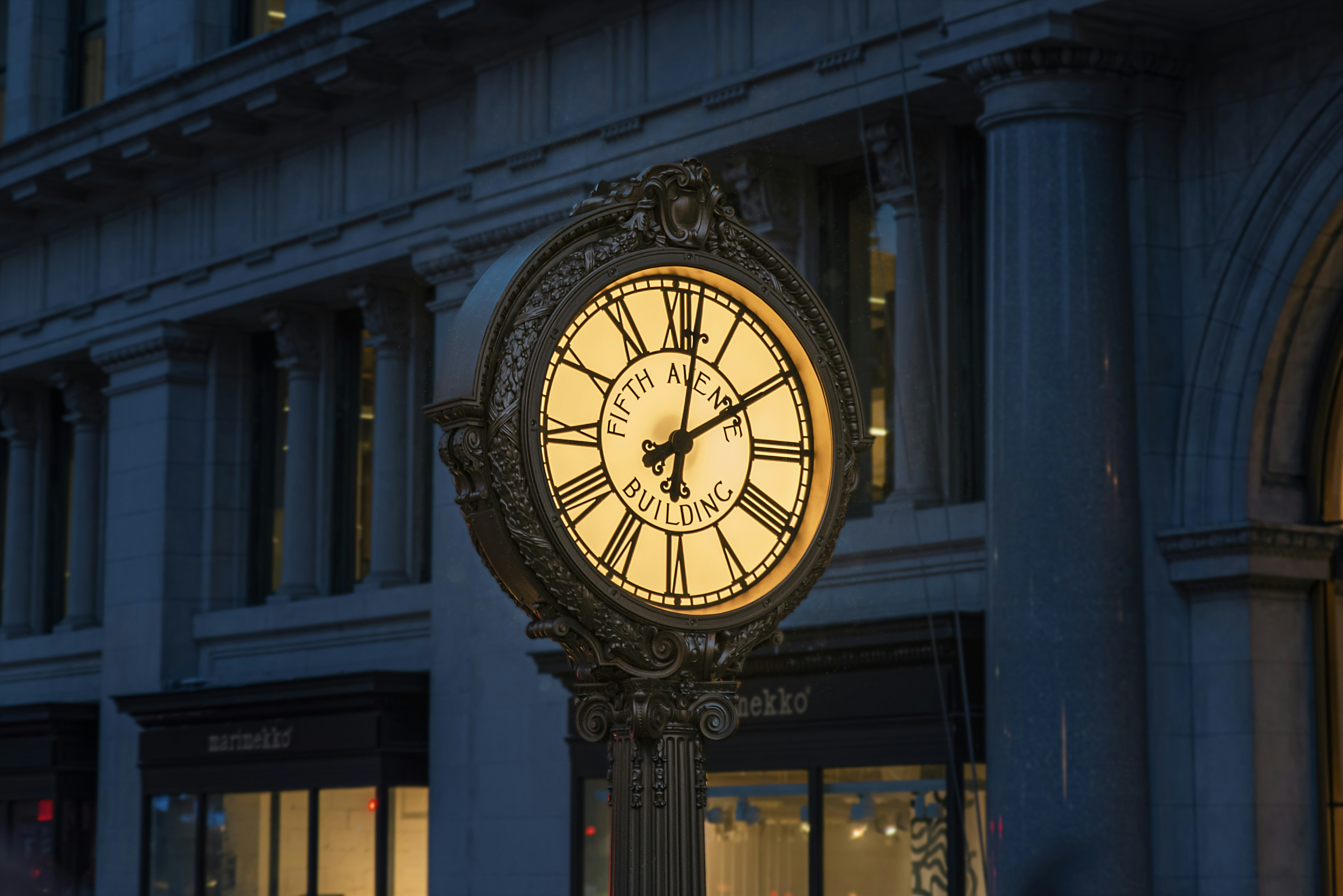 Illuminated clock featuring ornate design, showcasing the Fifth Avenue Building. The warm glow contrasts with the evening ambiance.