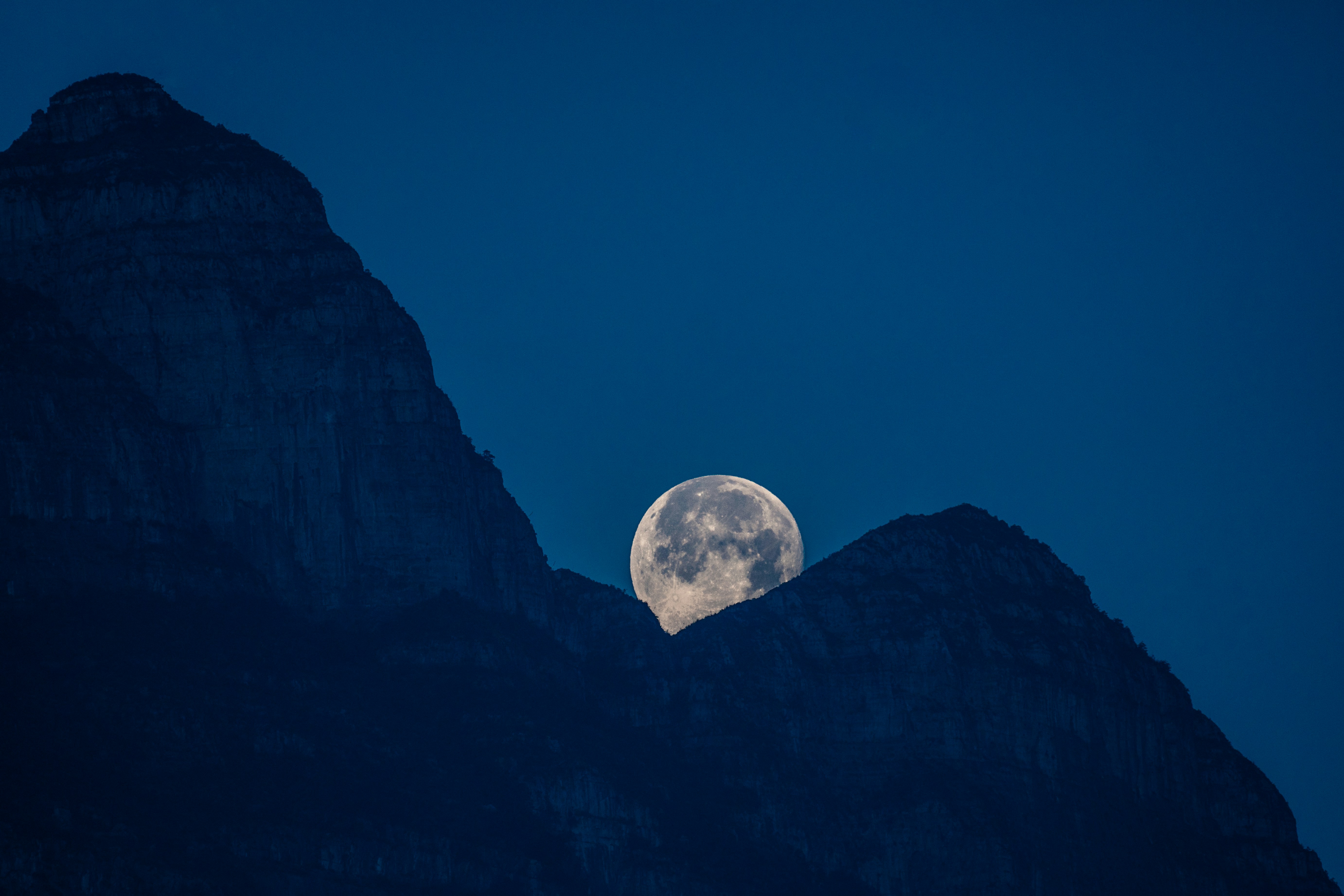 Full moon rising between two silhouetted mountain peaks against a deep blue sky.