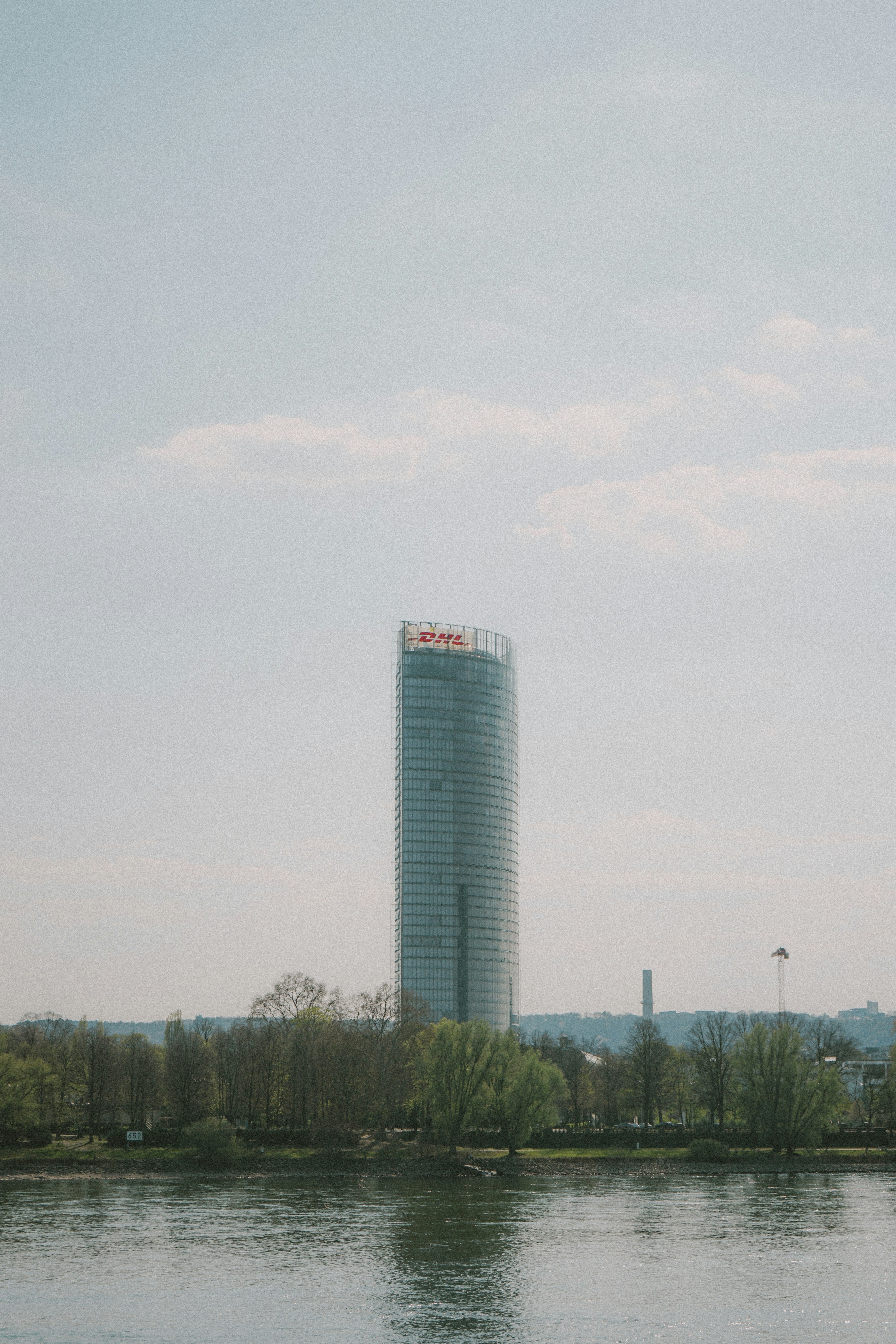 Modern glass tower rising beside a tranquil river, framed by lush greenery and a clear sky.