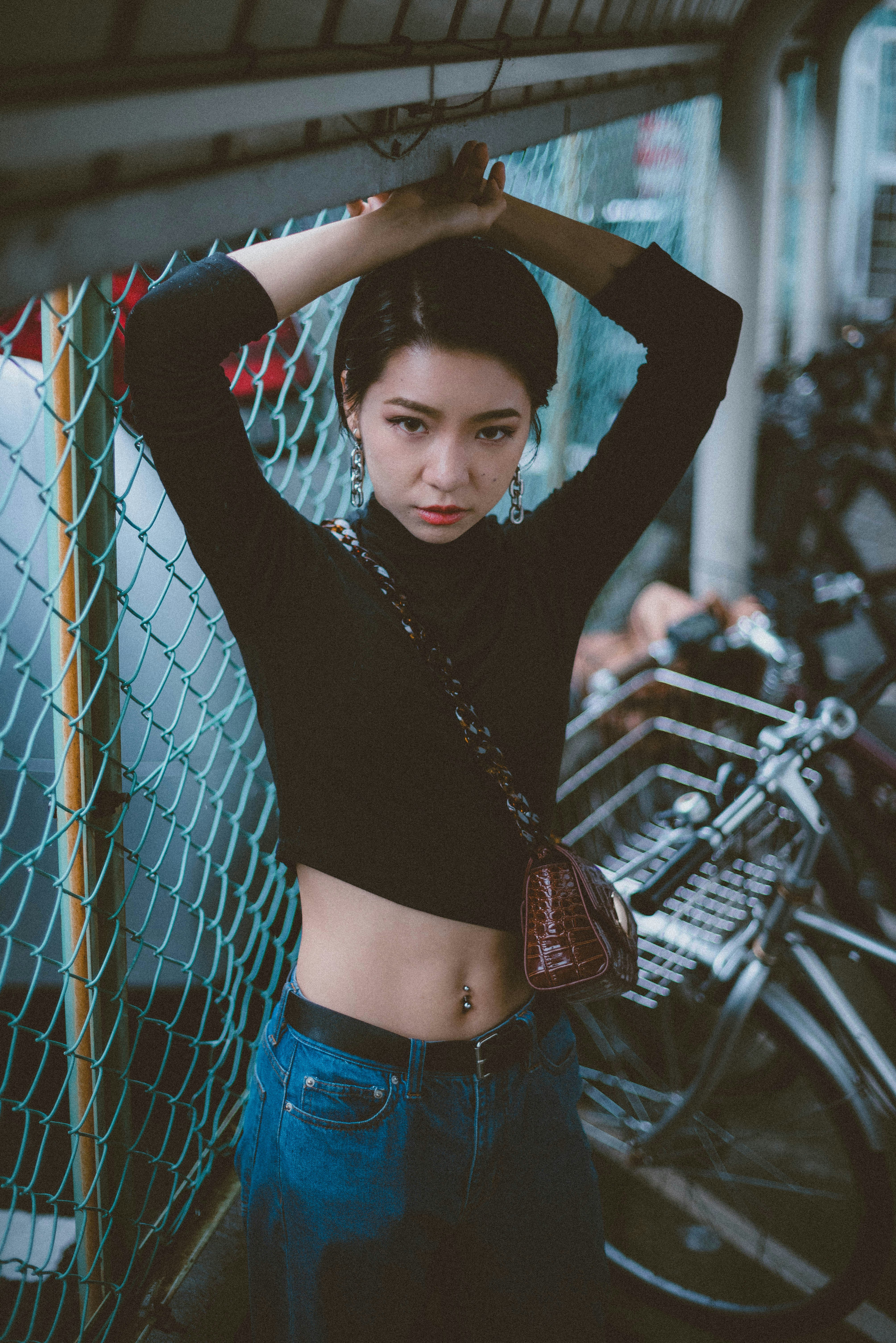 A young woman poses against a textured backdrop of a chain-link fence and bicycles, showcasing a blend of urban style and confidence.