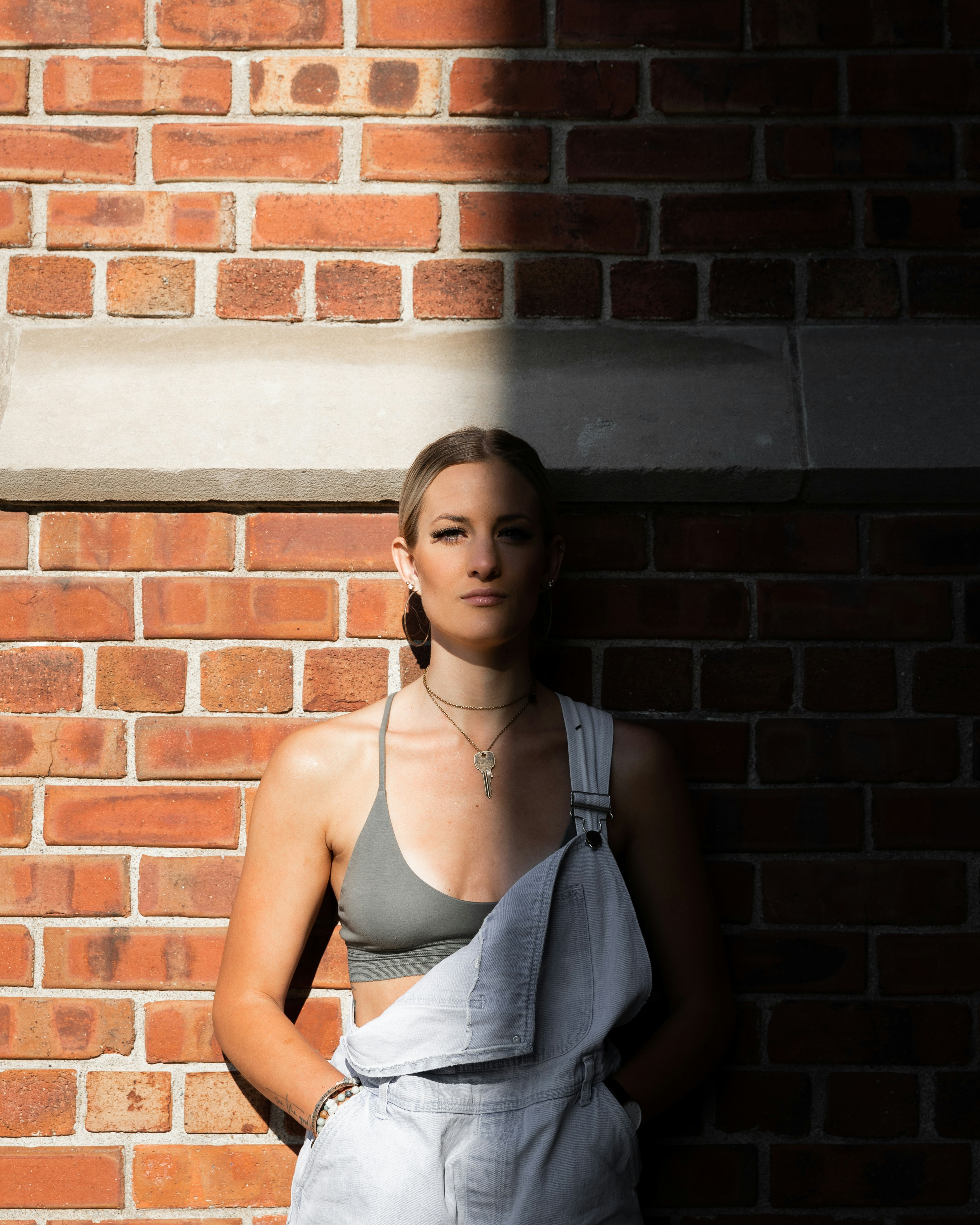 Woman in a gray top and white overalls leaning against a sunlit brick wall.