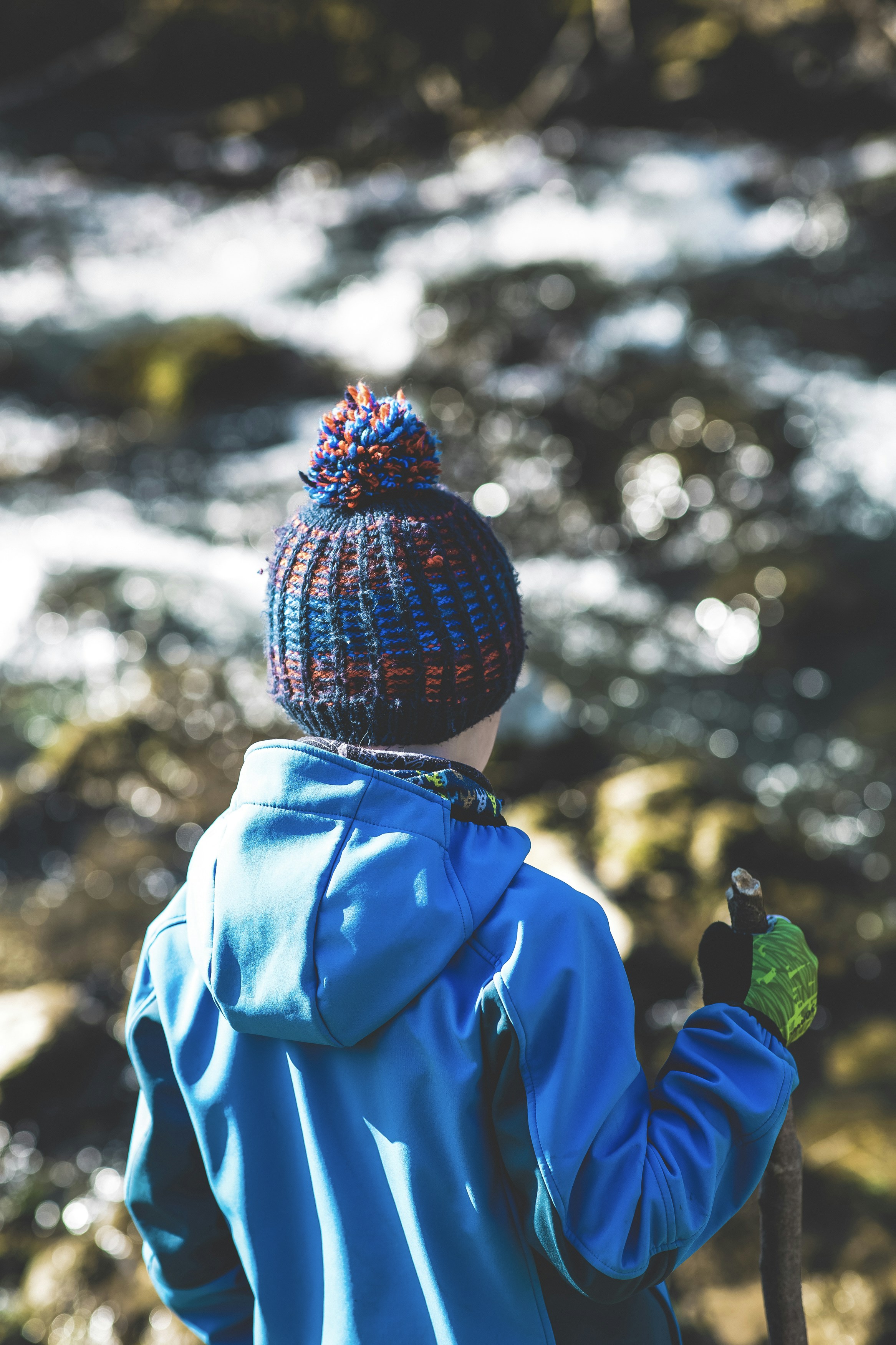 Child in a blue jacket and colorful hat gazing at a flowing stream, holding a stick. Sunlight glimmers on the water's surface.
