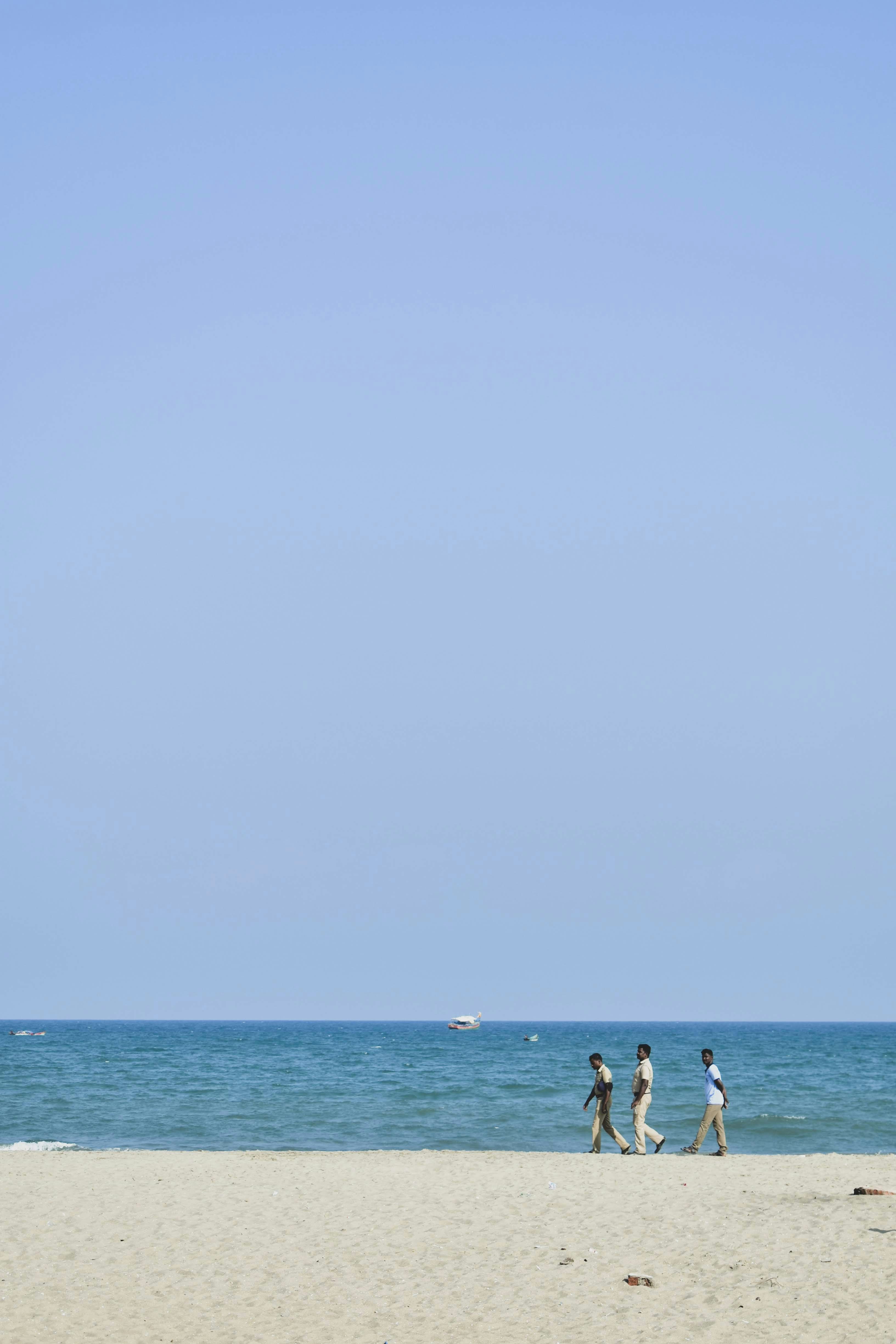 Three individuals walking along a sandy beach under a clear blue sky, with gentle waves lapping at the shore.