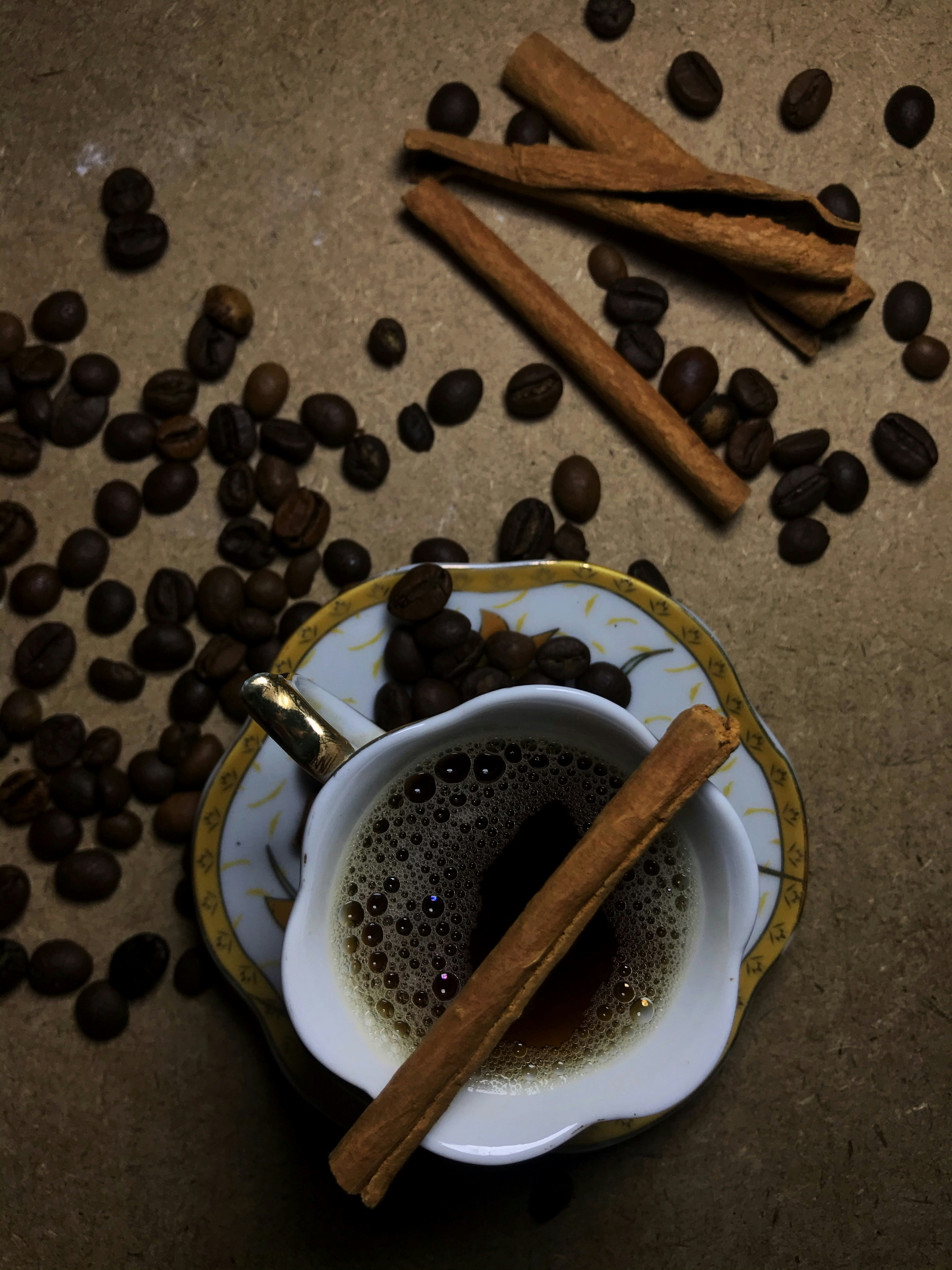 Delicate porcelain cup filled with dark coffee, garnished with a cinnamon stick, surrounded by scattered coffee beans.