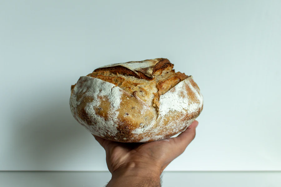 A hand holds a round rustic sourdough loaf with a crackled crust and flour dusting against a plain light background.