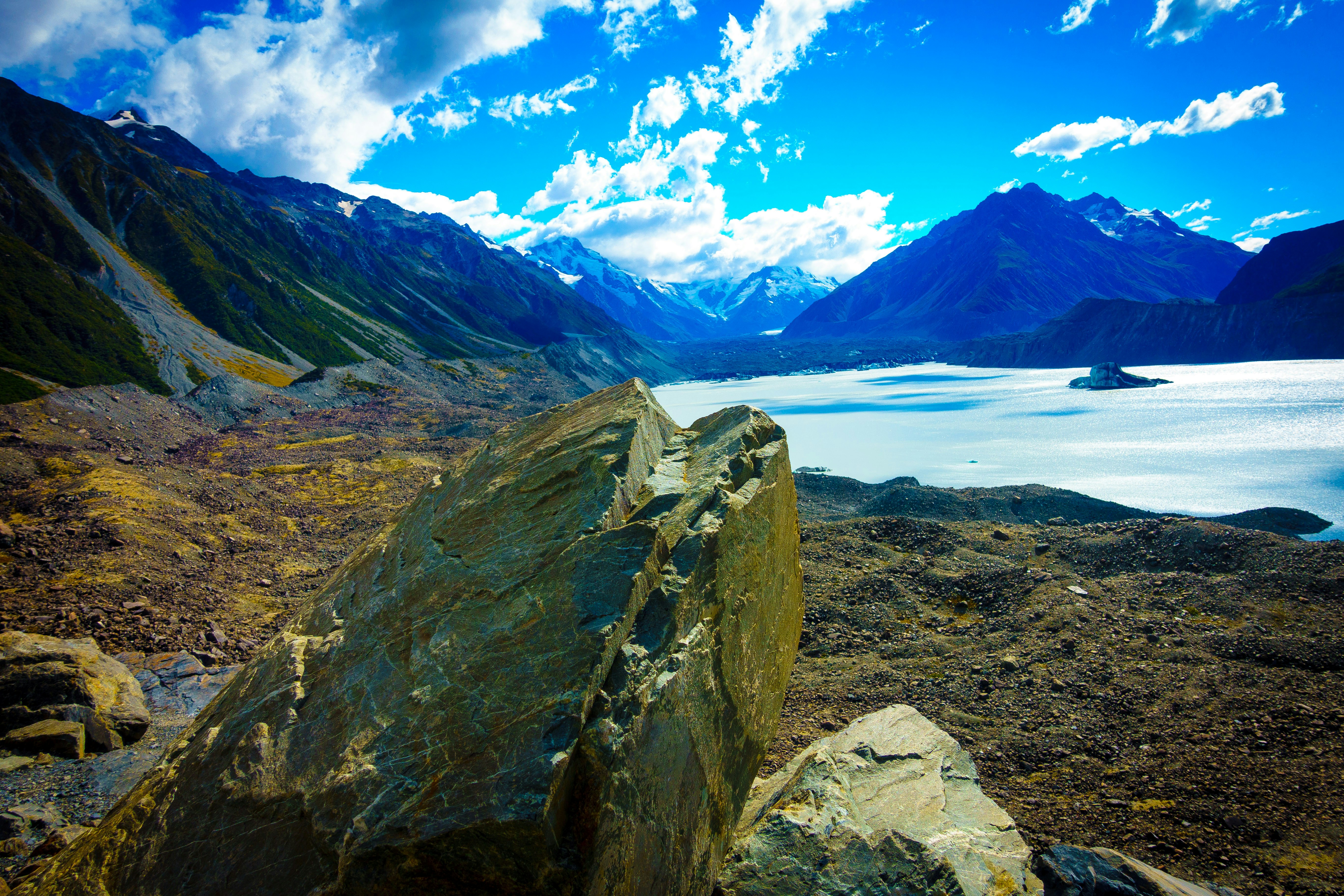 Jagged rocks overlook a glacial lake surrounded by towering alpine peaks under a vibrant blue sky.