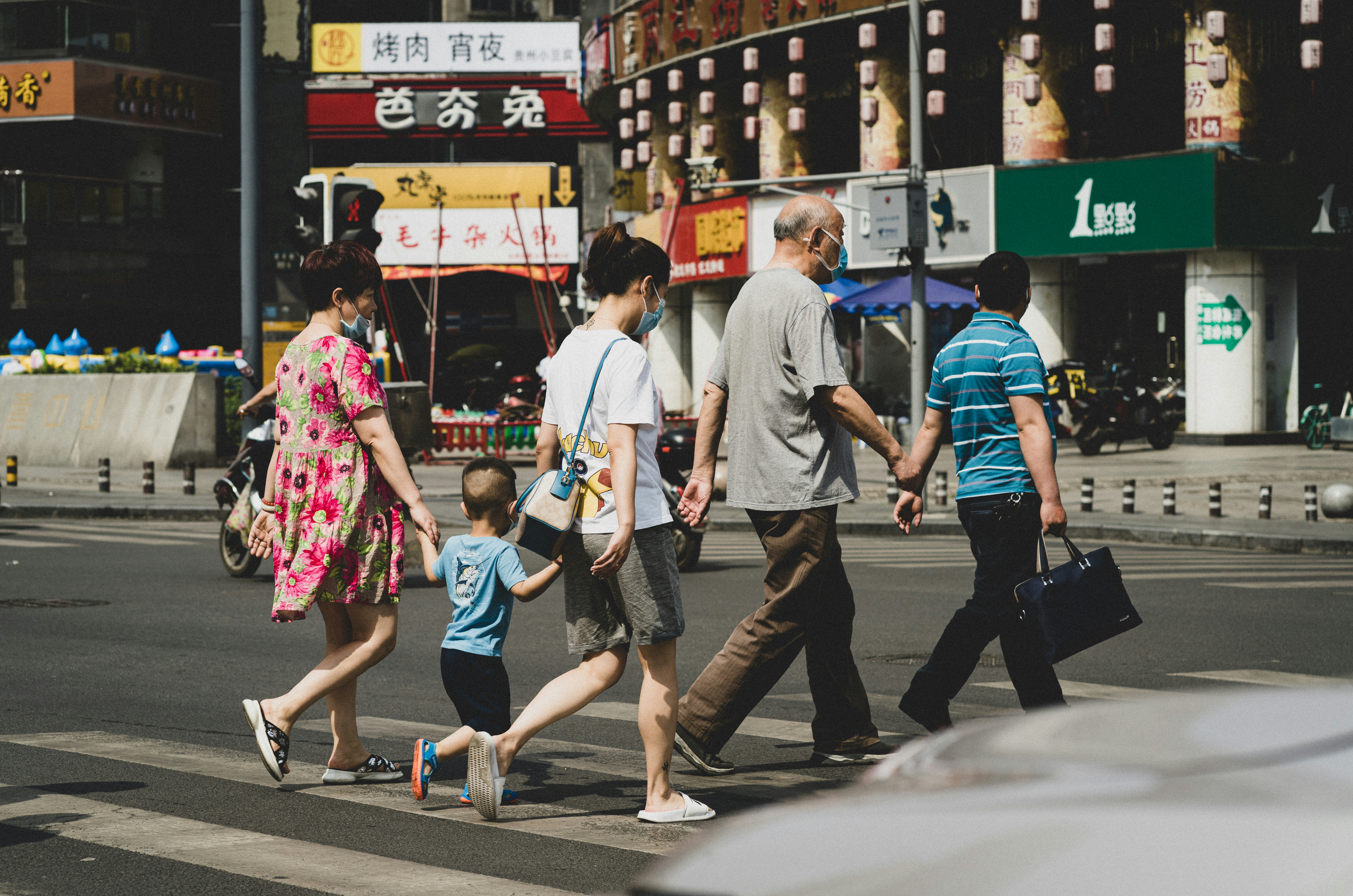 A family crossing a city street, with a child holding hands with an adult, set against a backdrop of vibrant storefronts and urban life.