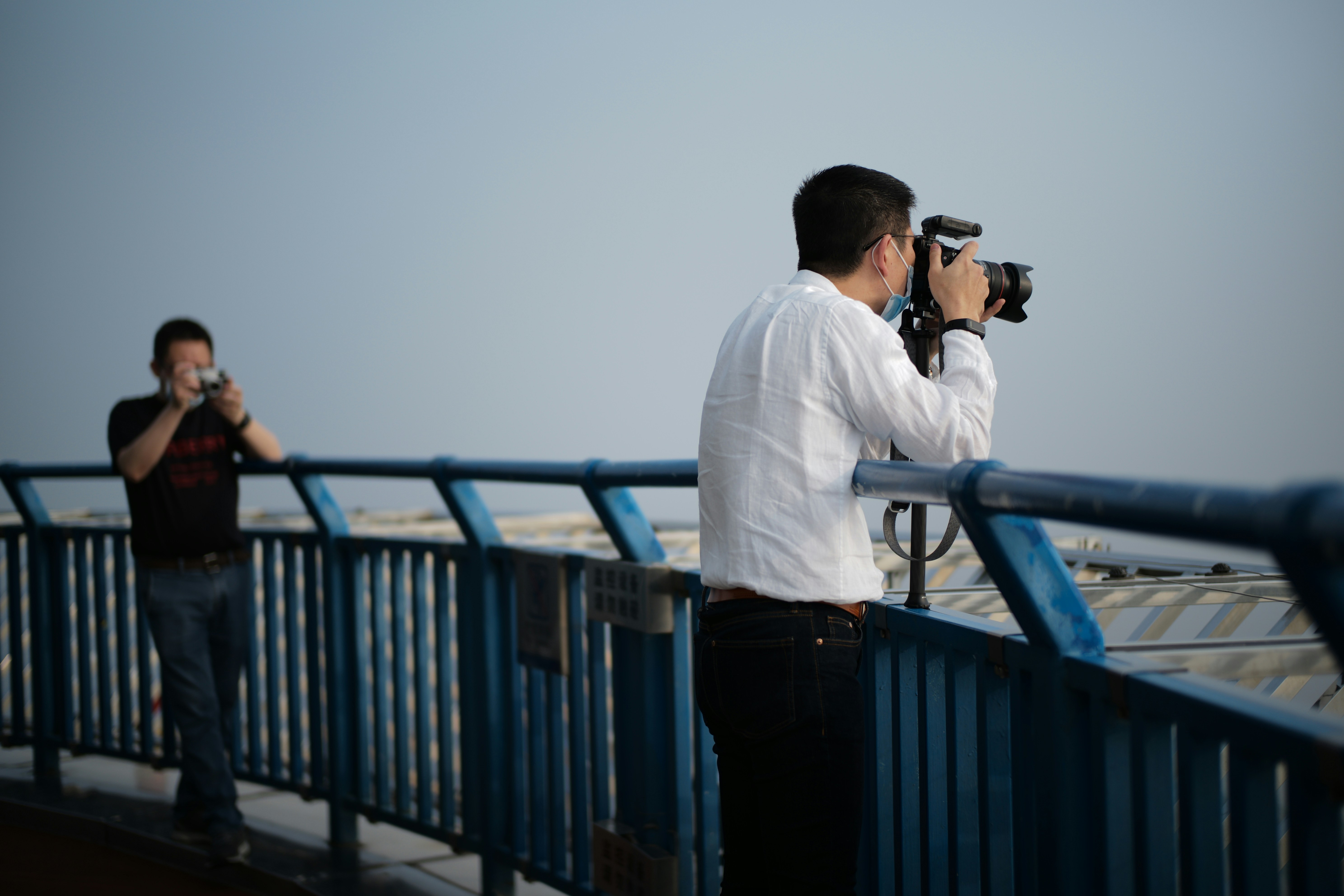 Two photographers on a balcony, one focused on capturing the view while the other poses with a camera. The scene highlights the passion for photography.