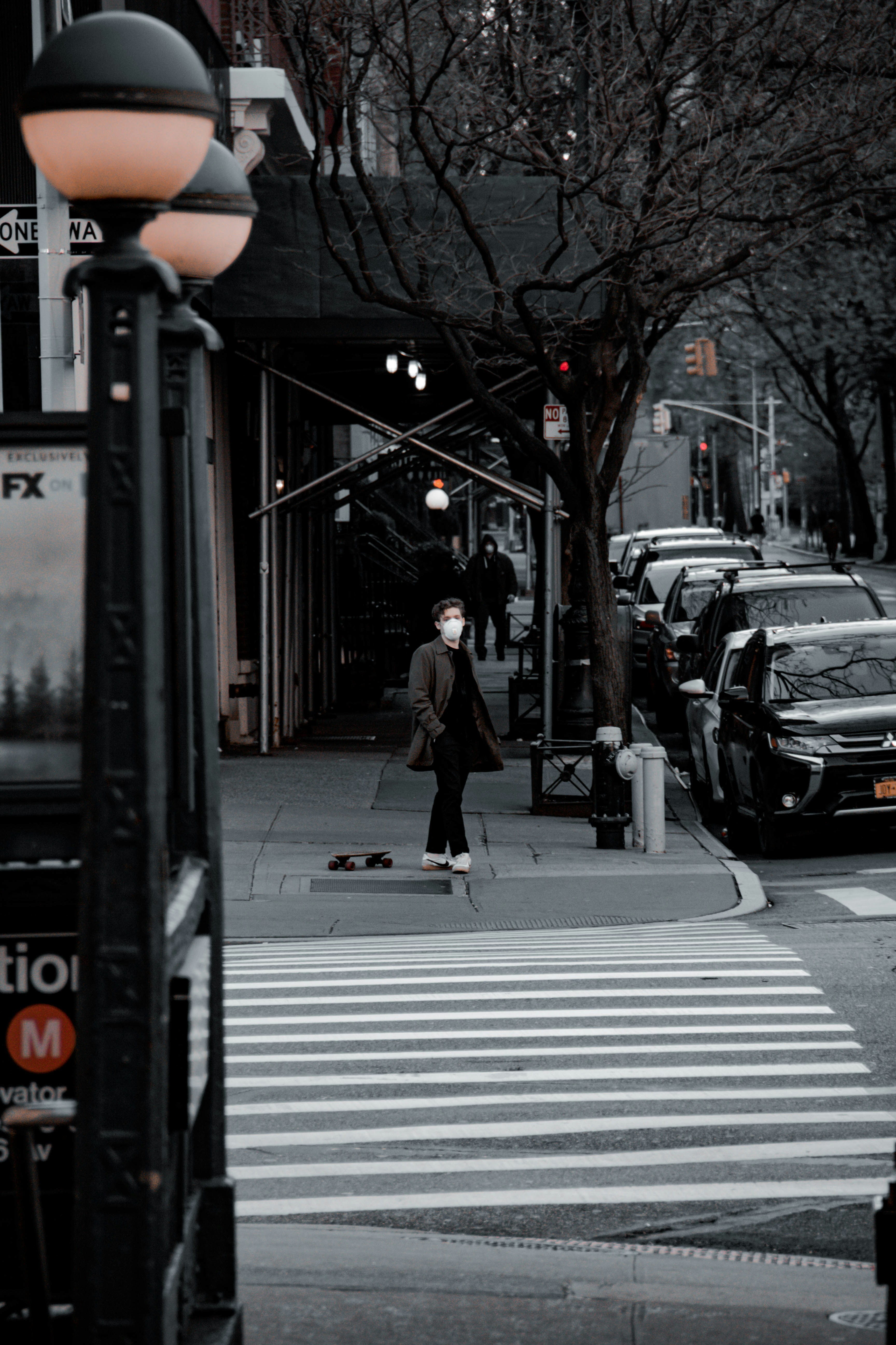 A person wearing a mask stands on a city street, surrounded by parked cars and bare trees, embodying a sense of isolation amidst urban life.
