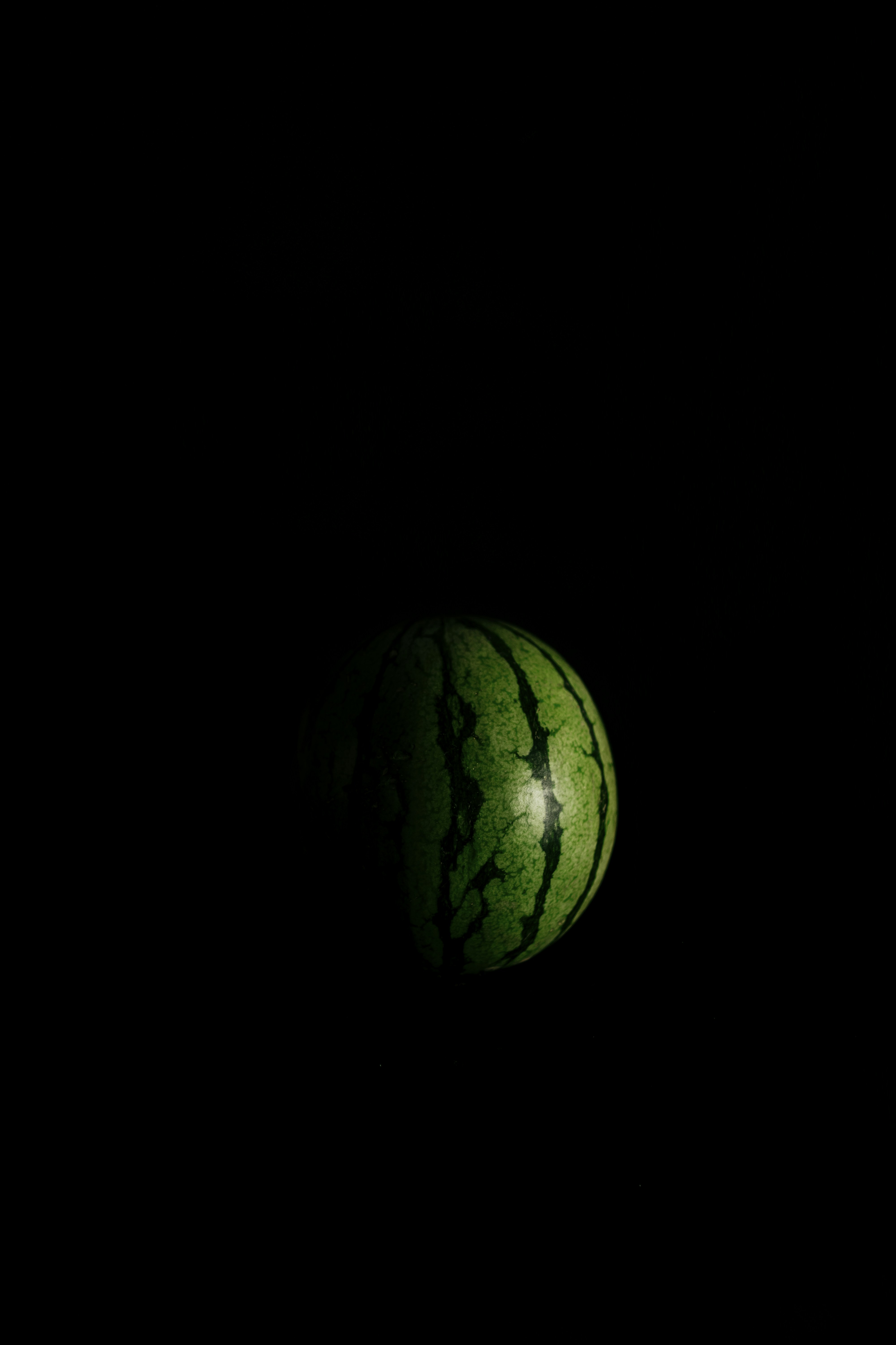A single watermelon illuminated against a dark backdrop, showcasing its vibrant green stripes and glossy surface.