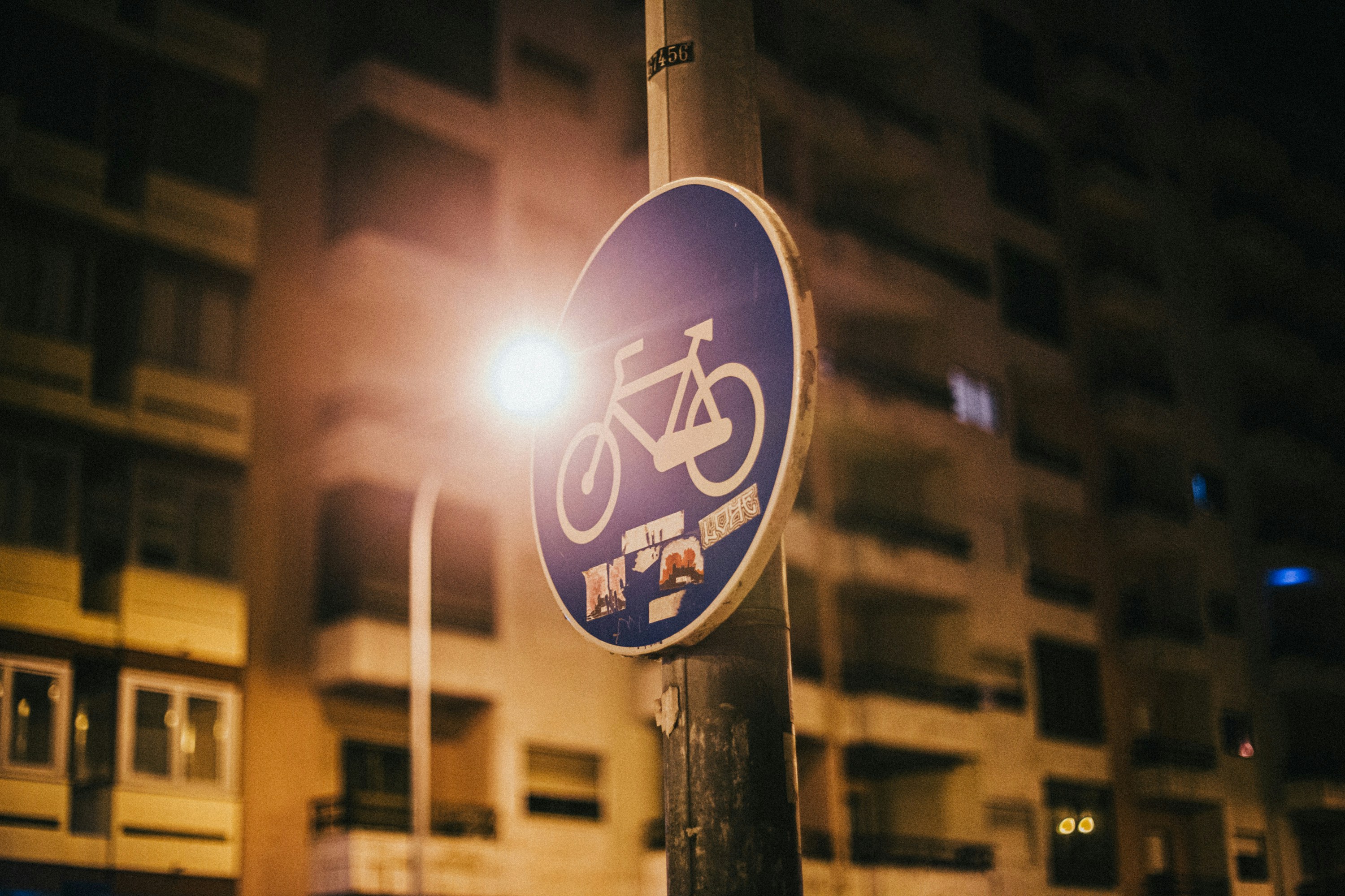 Bicycle sign illuminated by streetlight against a backdrop of urban architecture at night.