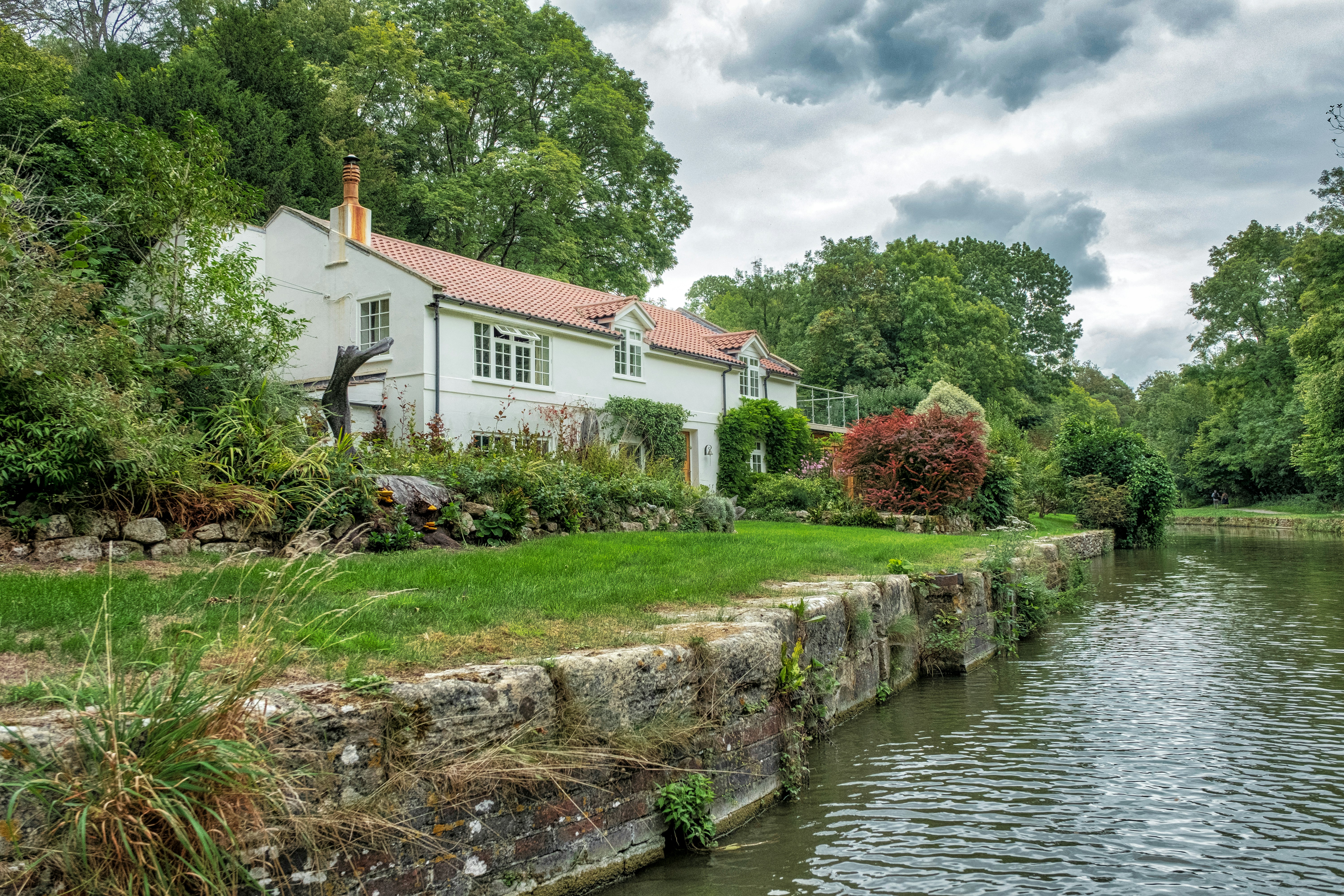 Charming white cottage with red roof nestled by a lush canal under a cloudy sky.