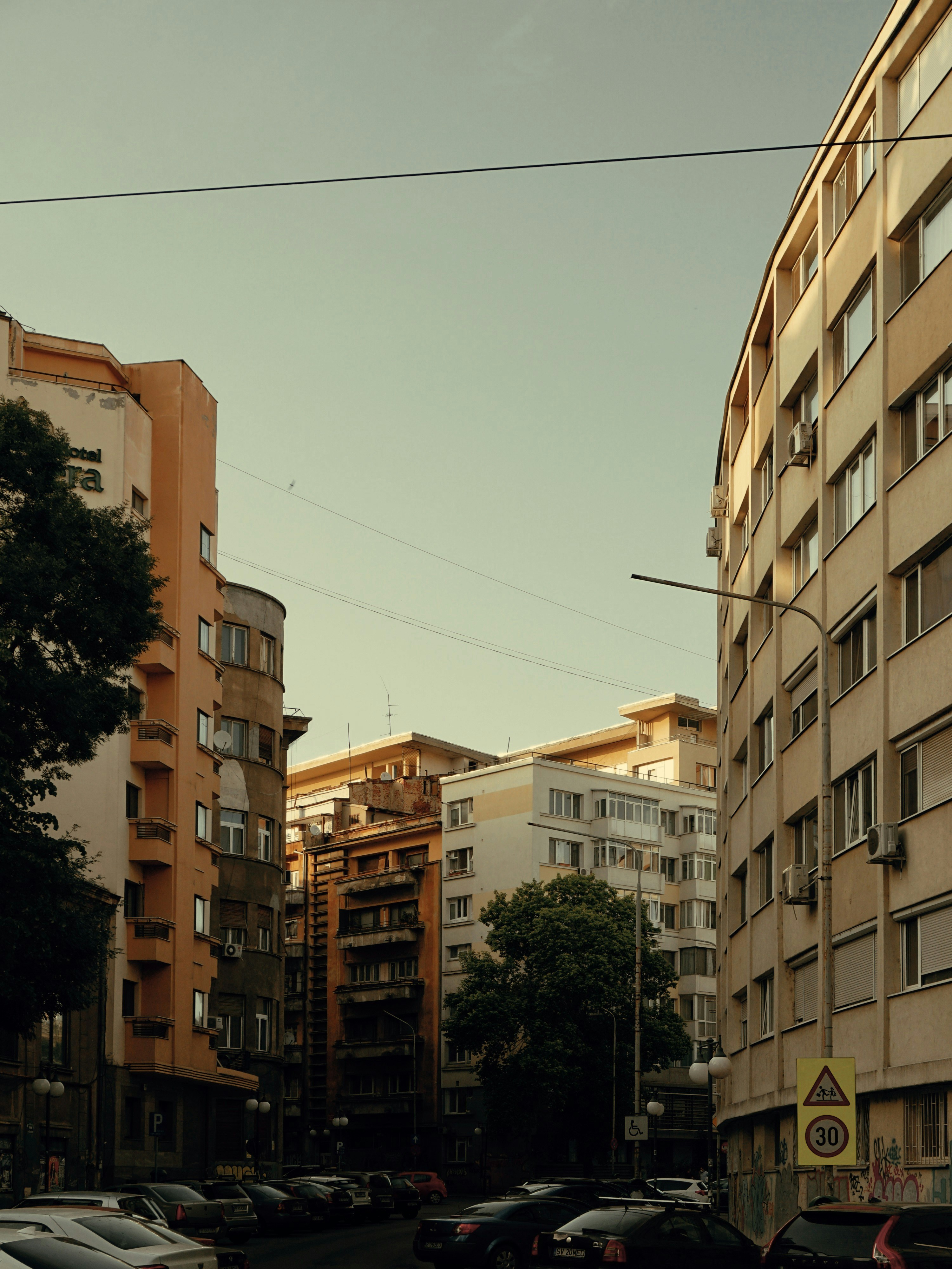 Narrow city street flanked by a mix of modern and vintage architecture, with parked cars and trees lining the road.