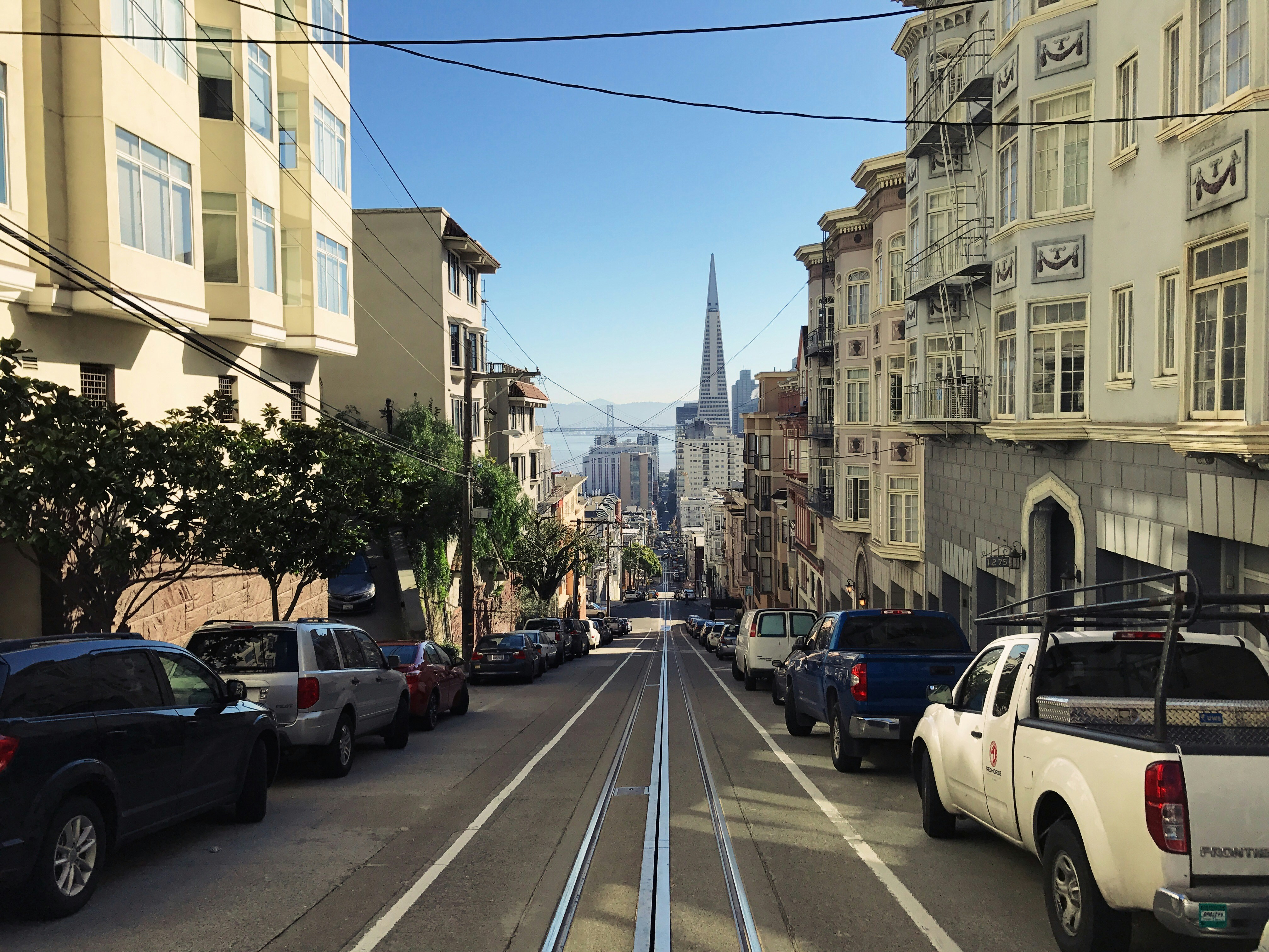 View down a steep San Francisco street lined with parked cars, leading towards the iconic Transamerica Pyramid in the distance.