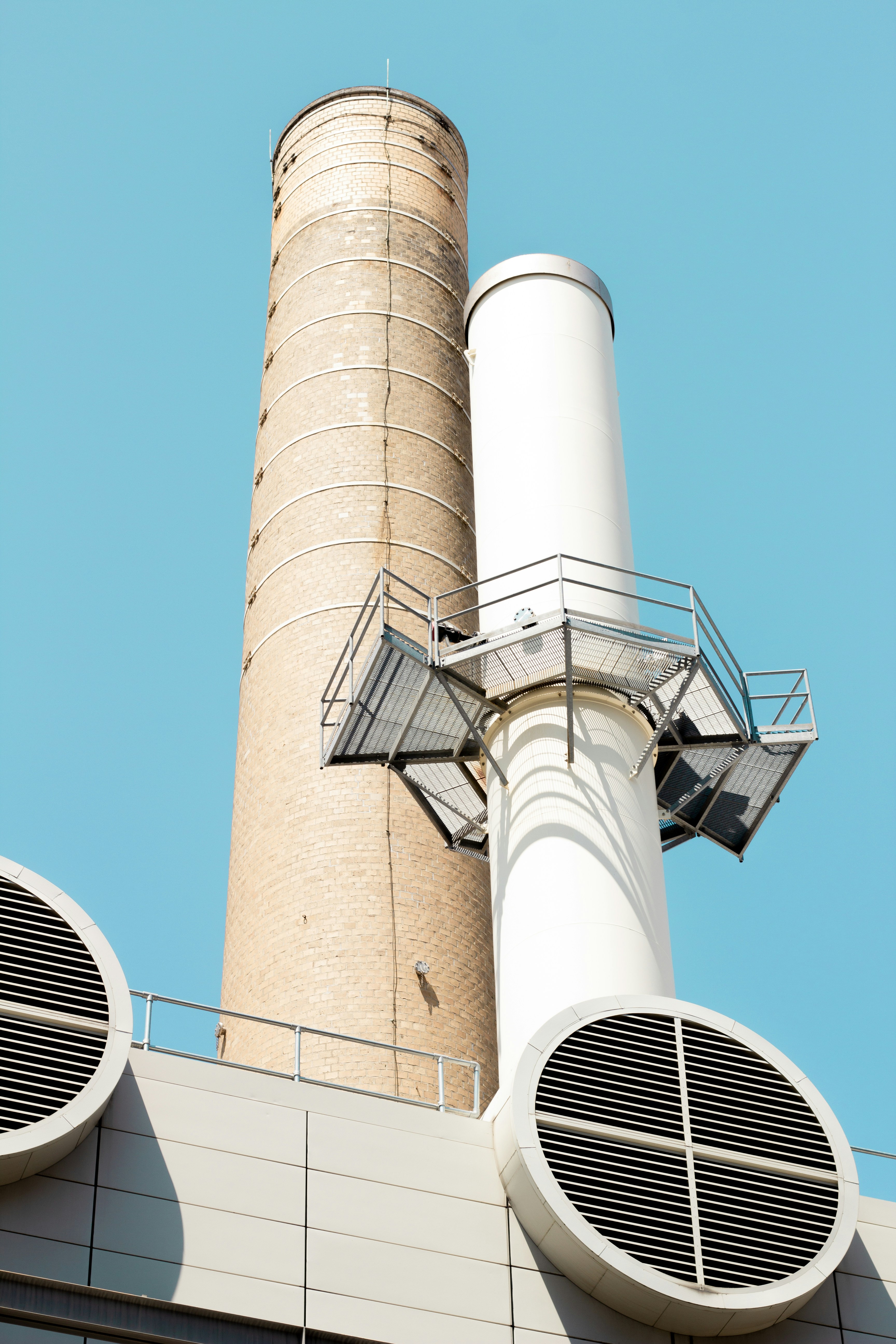 Two towering smokestacks rise against a clear blue sky, showcasing industrial architecture with a modern twist. The scene emphasizes the contrast between the structures and the vibrant backdrop.