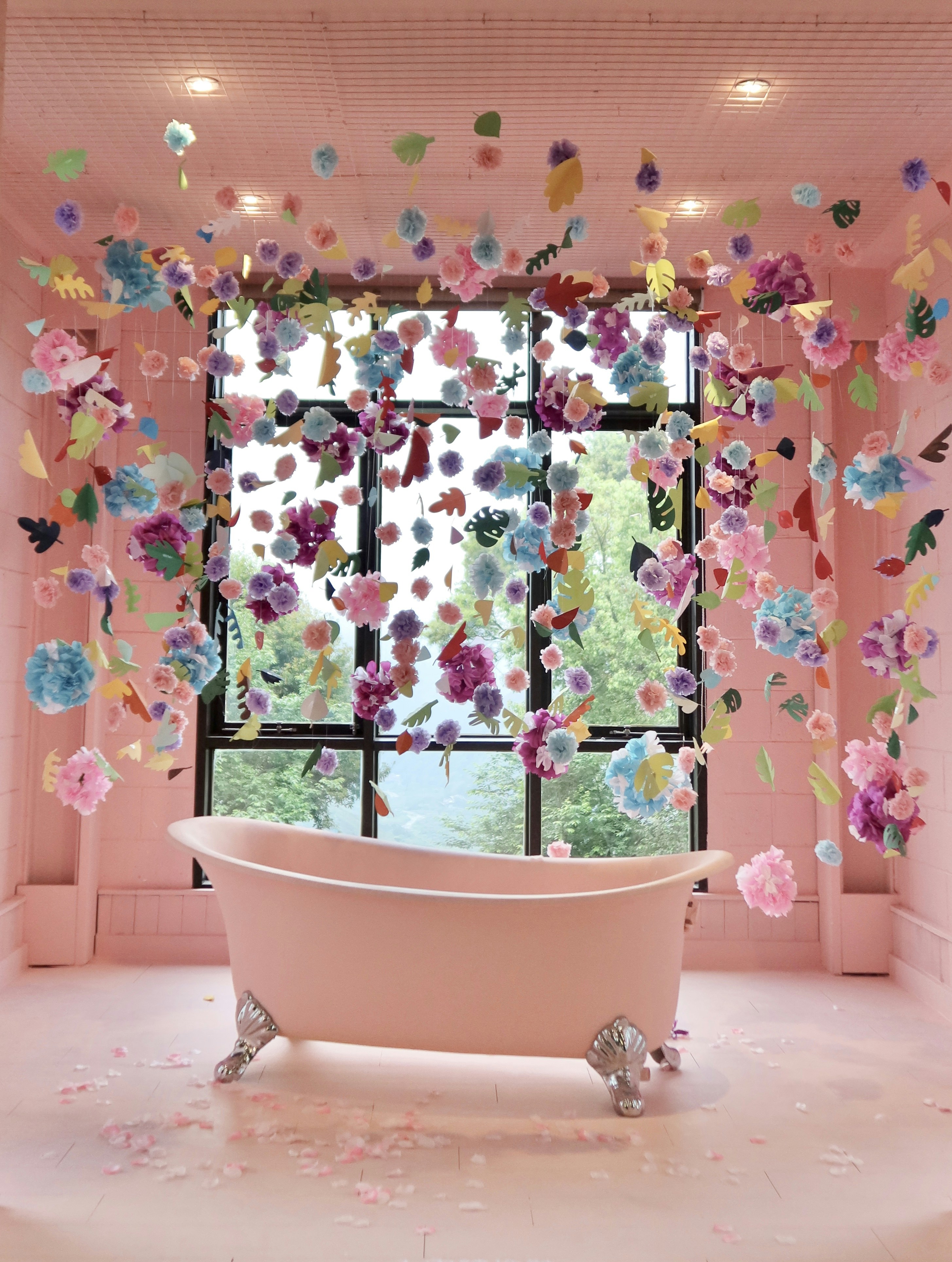 A vintage clawfoot bathtub sits in a pastel pink room adorned with vibrant hanging flowers and leaves, creating a serene and playful ambiance.