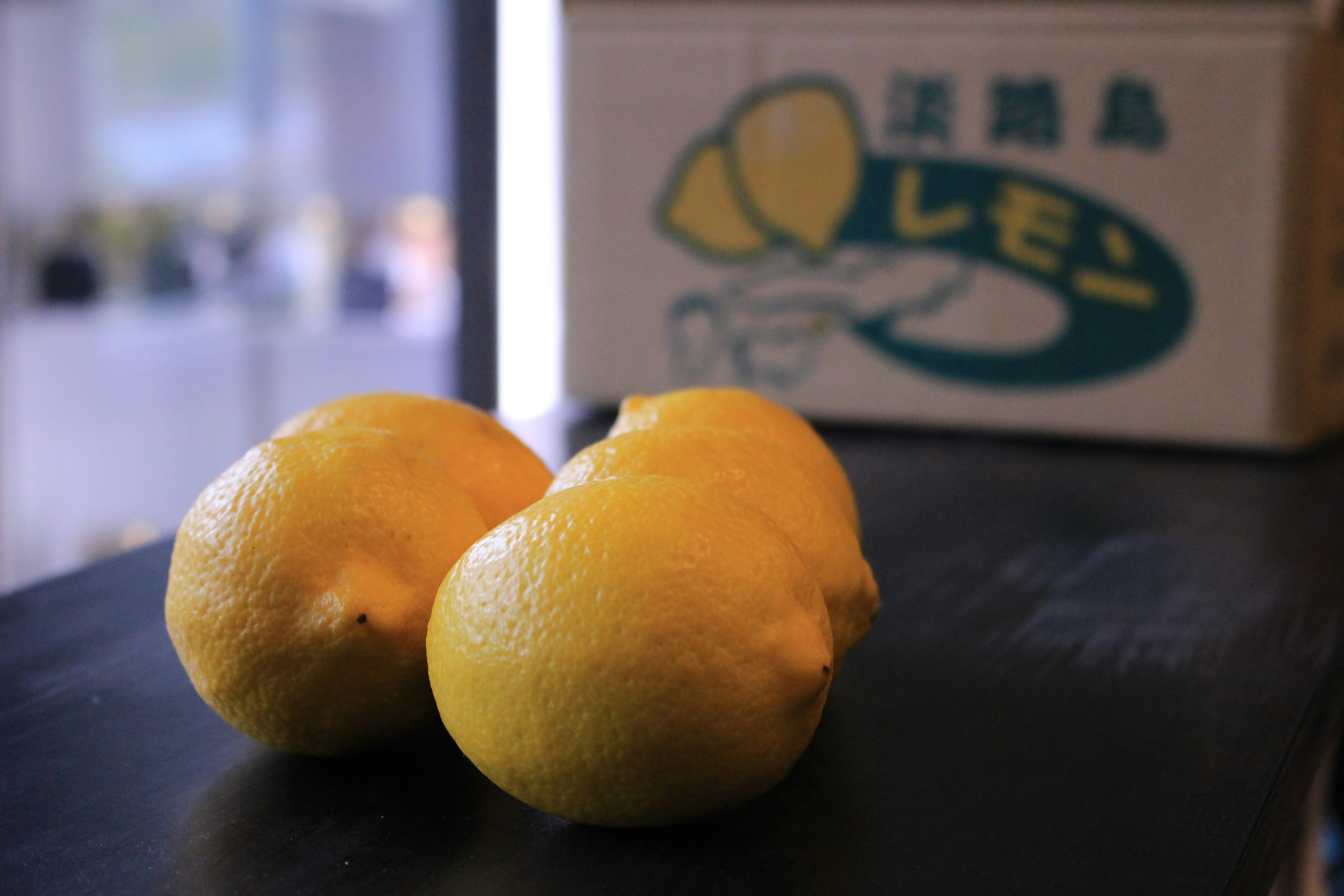 Four ripe lemons arranged on a dark surface with a box featuring a lemon graphic in the background.
