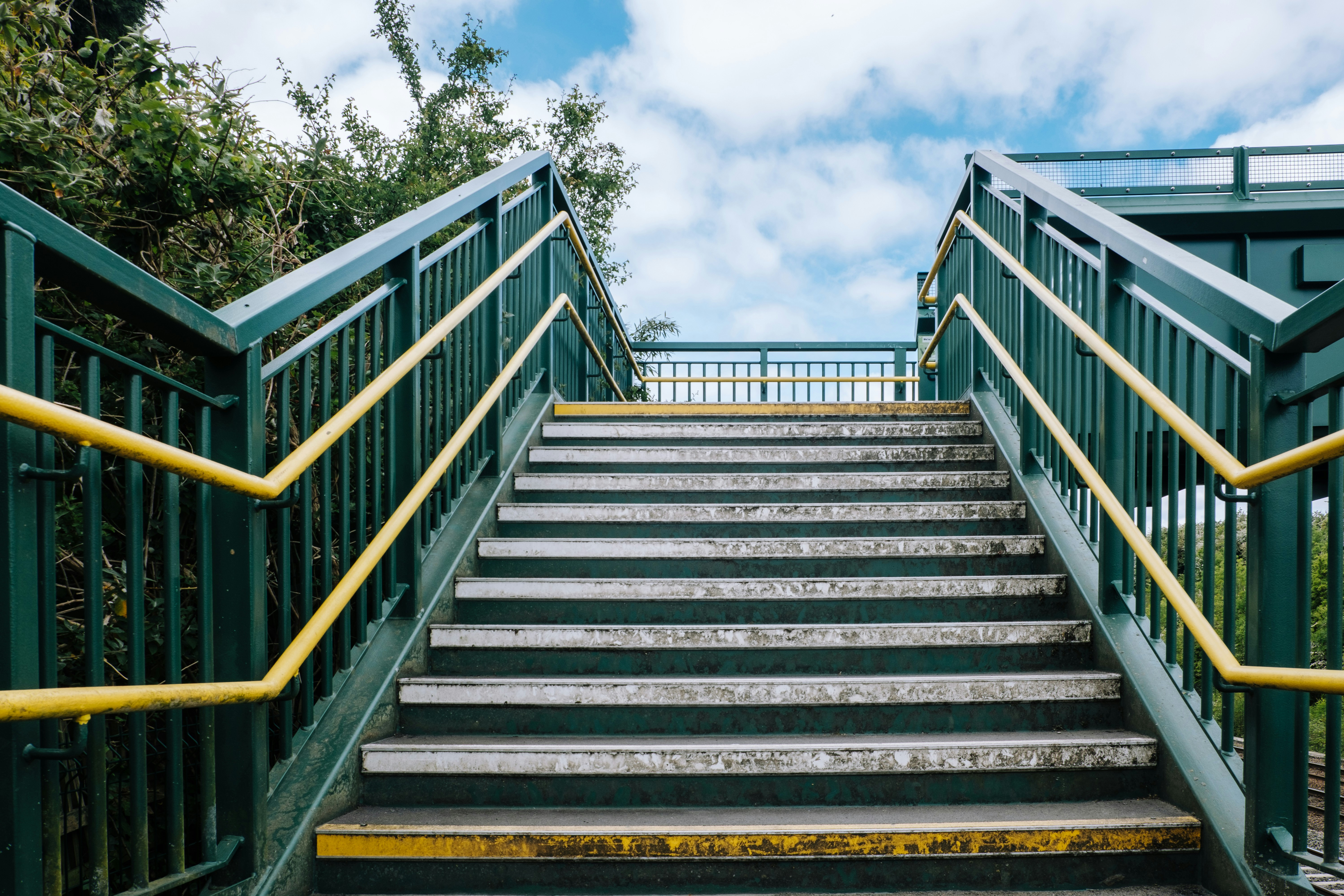 Green metal staircase leading upward, flanked by railings and surrounded by foliage under a cloudy sky.