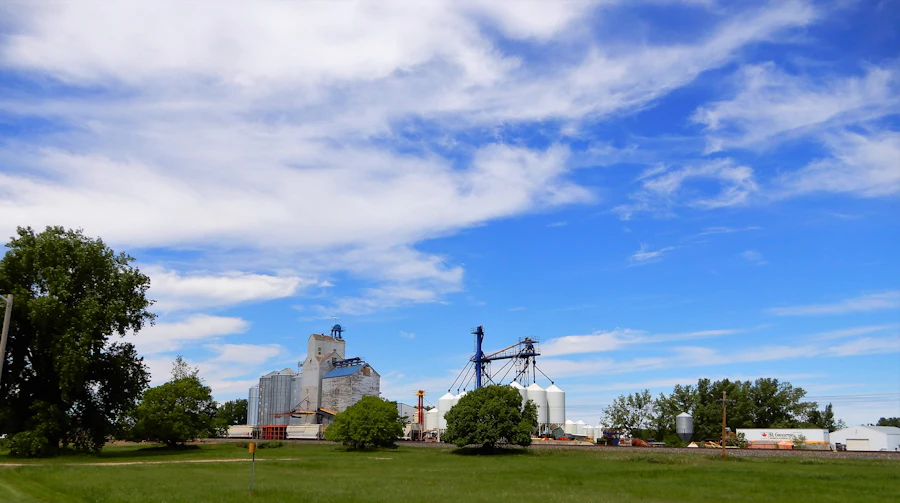 Wide prairie foreground with green grass and trees framing distant grain silos and industrial structures. A bright blue sky with scattered clouds dominates the scene.