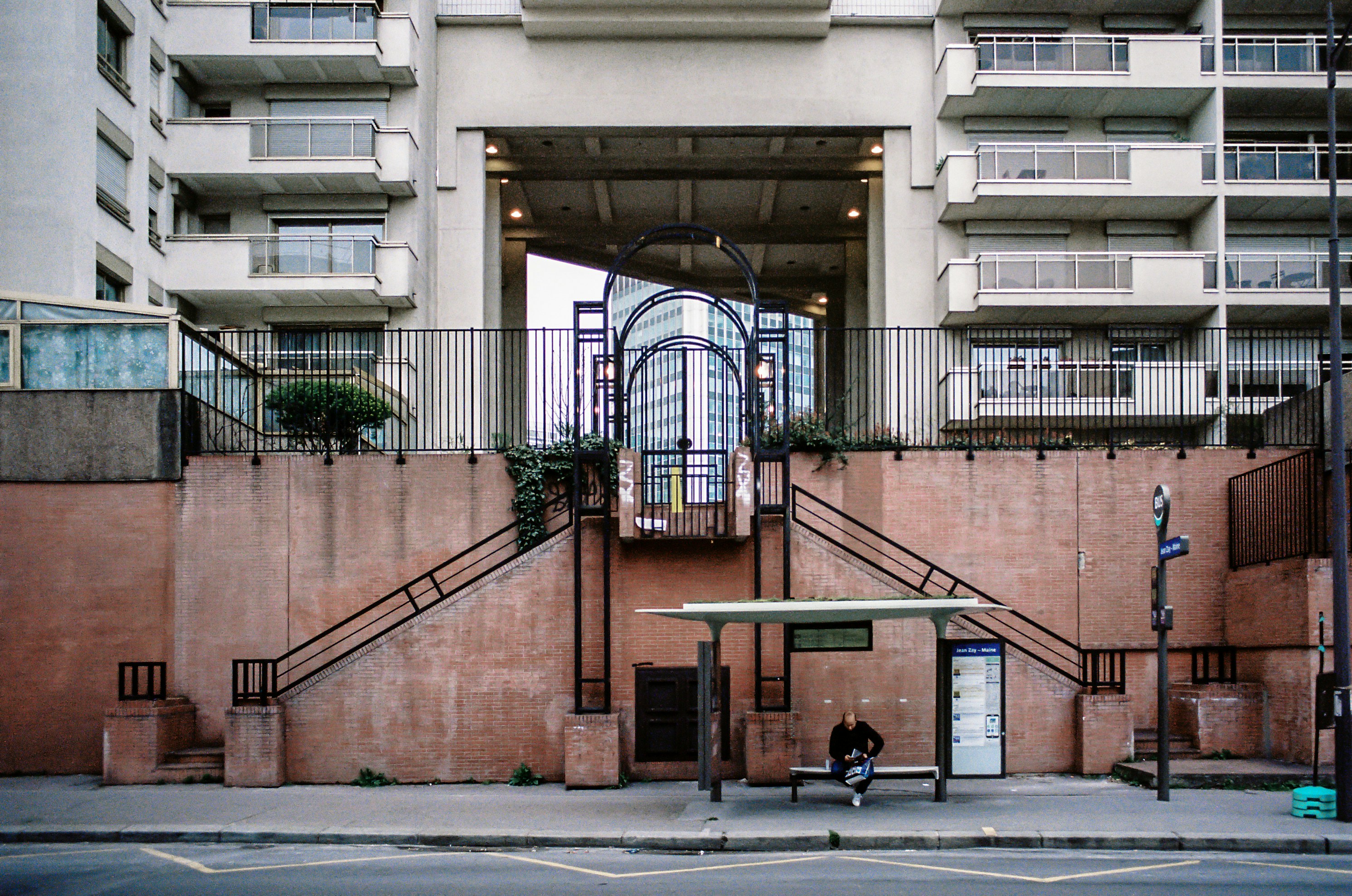 A solitary figure waits at a bus stop beneath a modern architectural overpass, framed by stark concrete and angular lines.