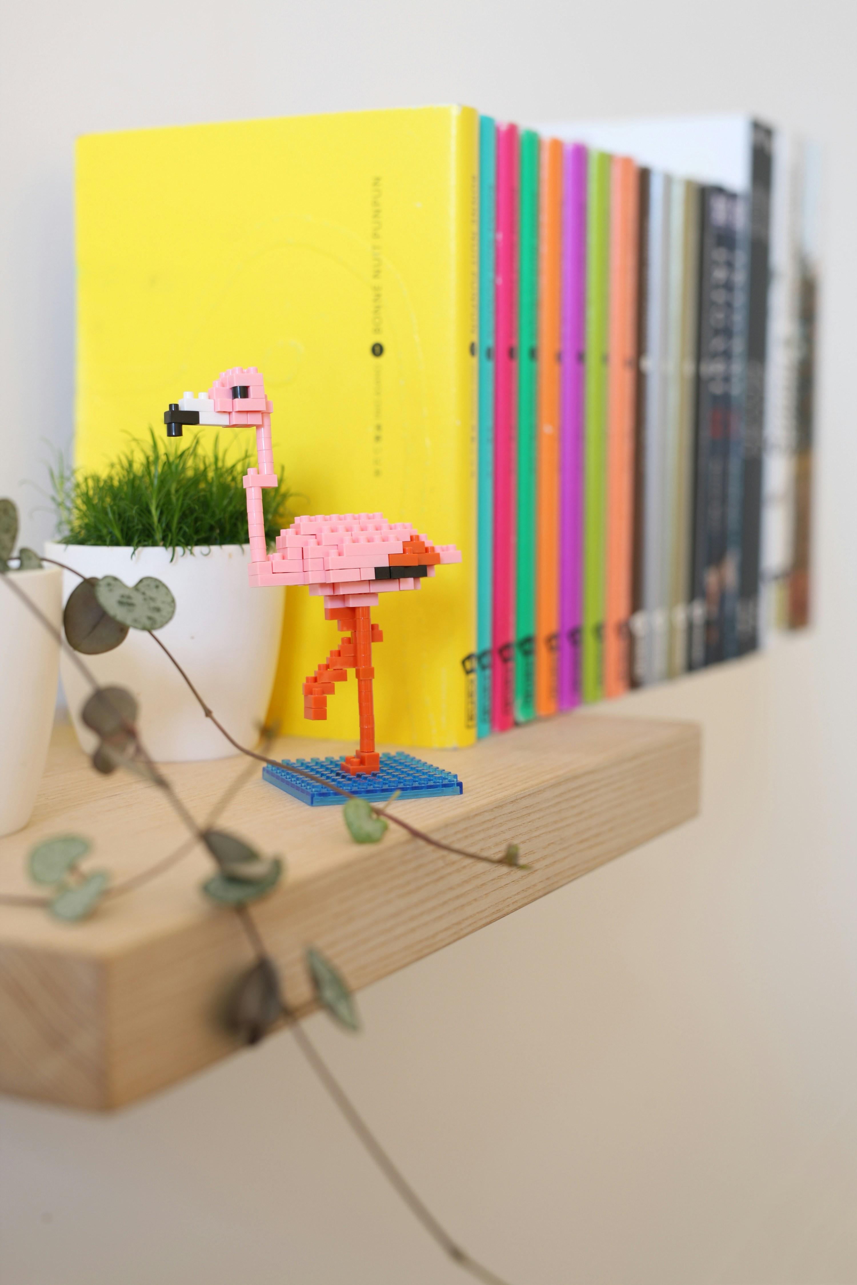Pink Lego flamingo figurine sits on a blue base beside a white plant pot, in front of a row of brightly colored book spines on a wooden shelf.