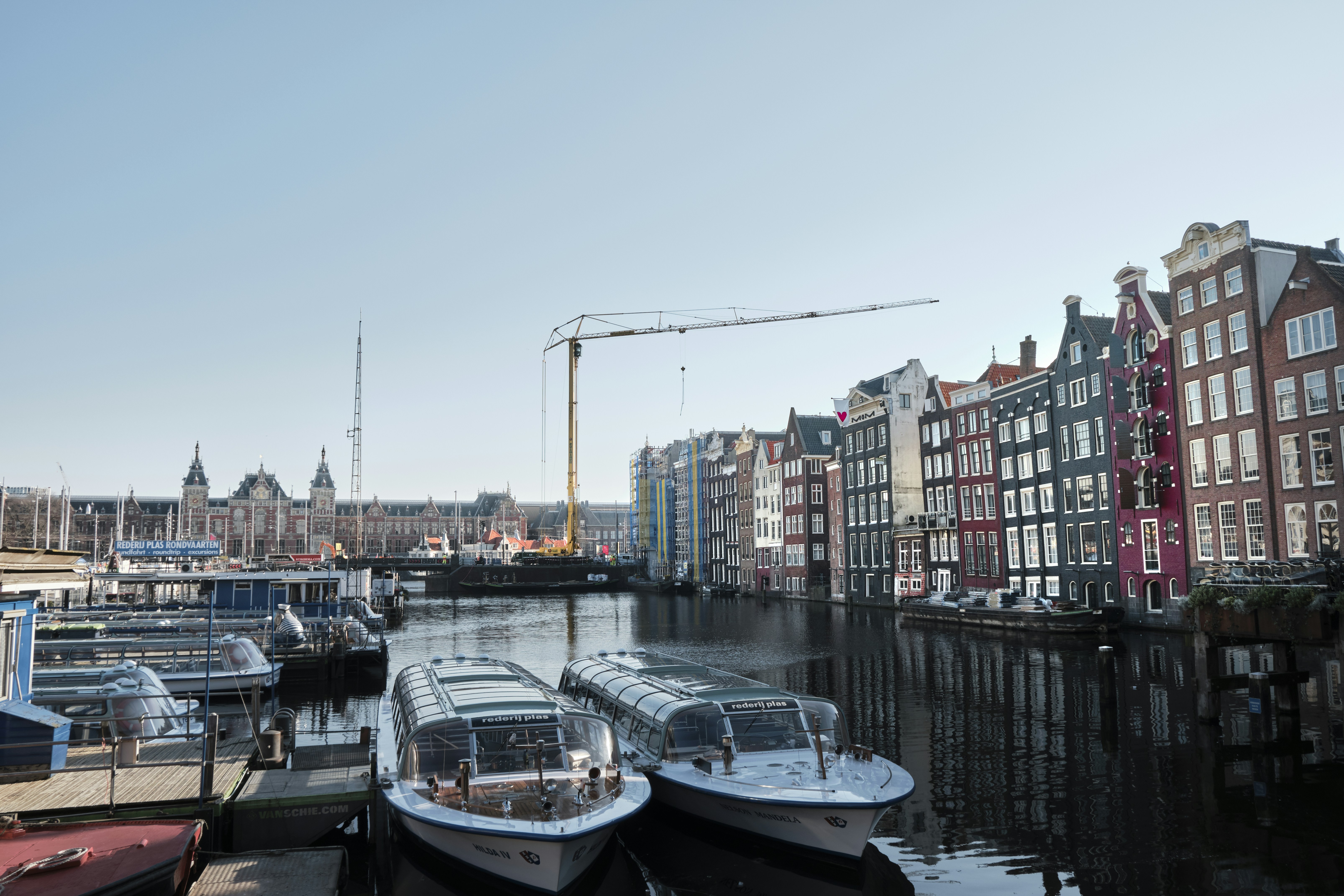 Colorful canal houses line the waterfront, with boats docked in the foreground and a crane in the background. The scene captures the essence of Amsterdam's vibrant urban landscape.
