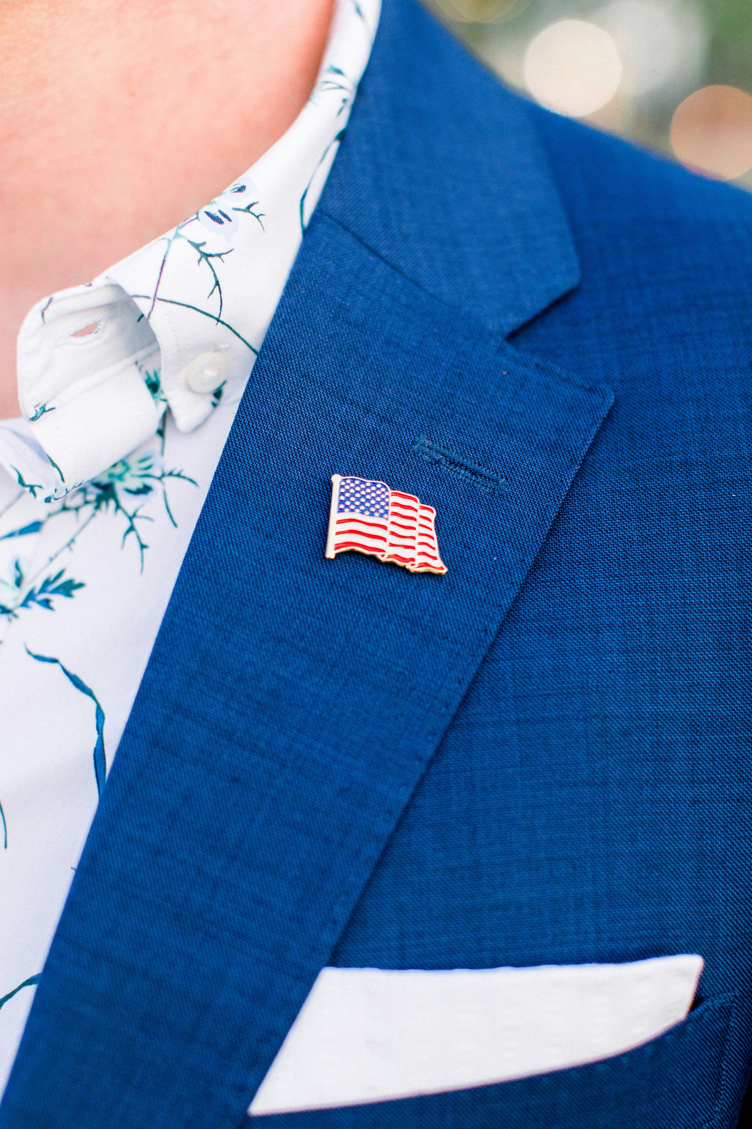 Close-up of a lapel pin featuring the American flag, worn on a blue blazer with a floral-patterned shirt beneath.