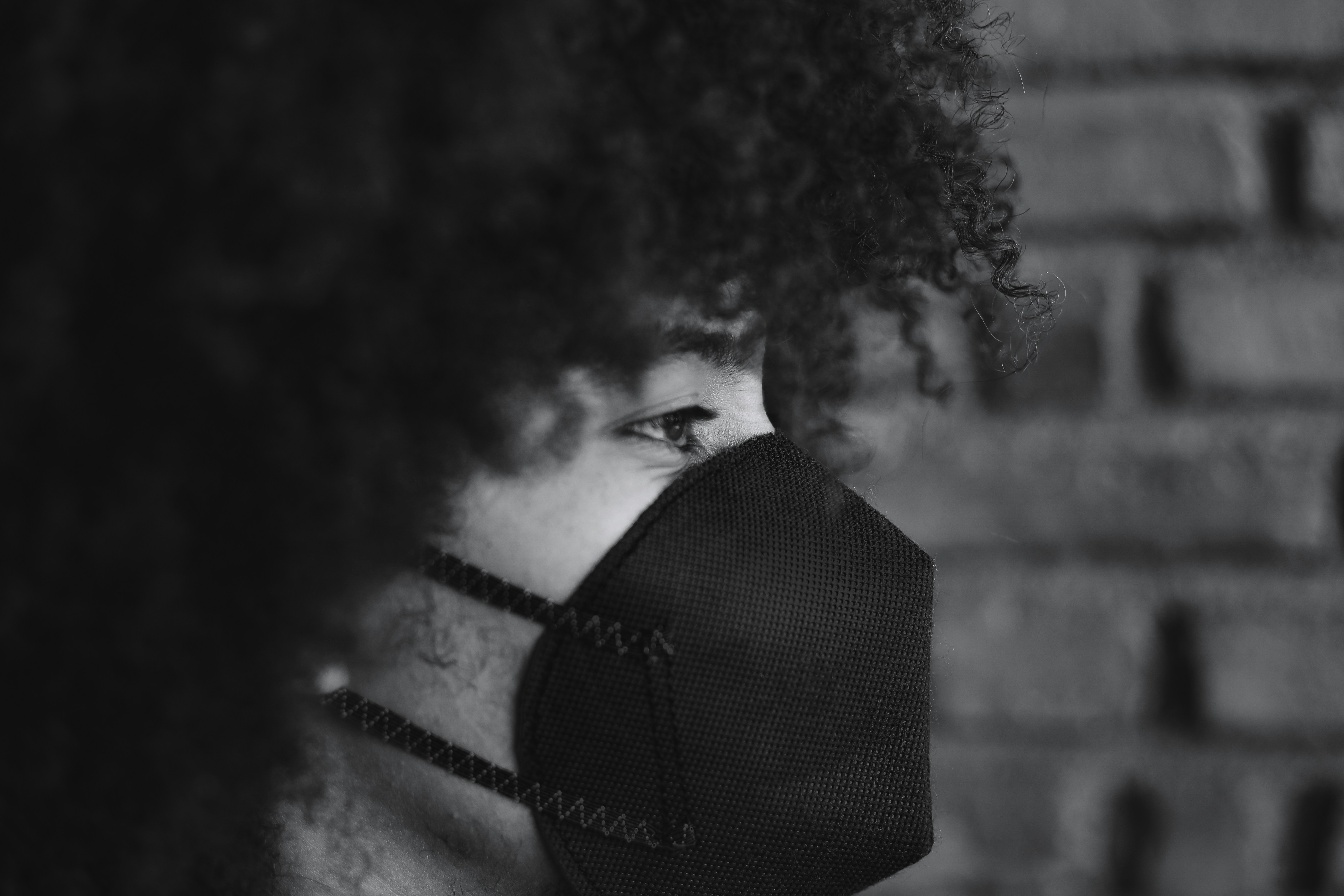 Close-up of a person wearing a black mask, highlighting their thoughtful expression against a textured brick background.