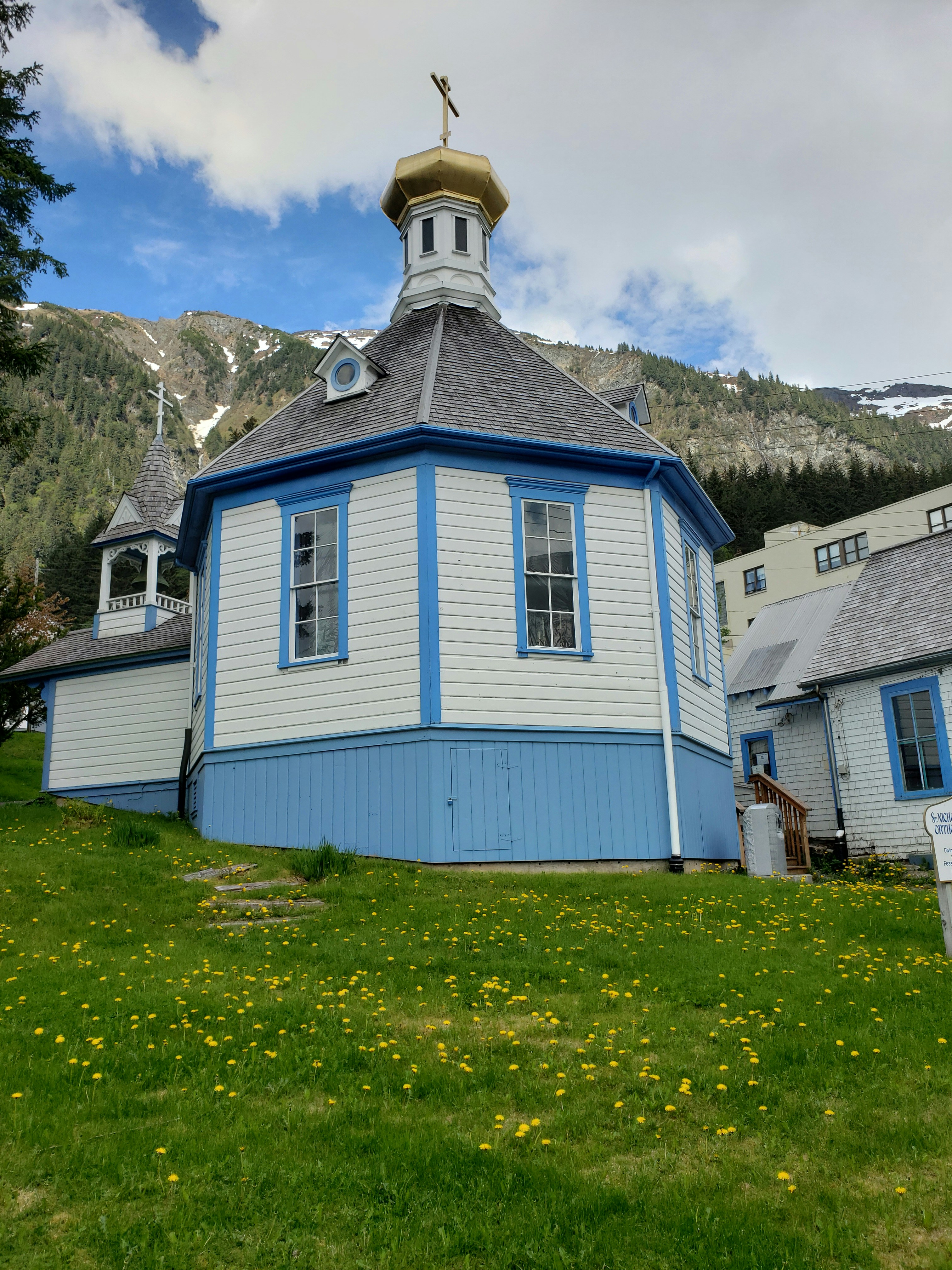 A charming octagonal church with a golden cross atop, surrounded by lush green grass and dandelions, set against a mountainous backdrop.