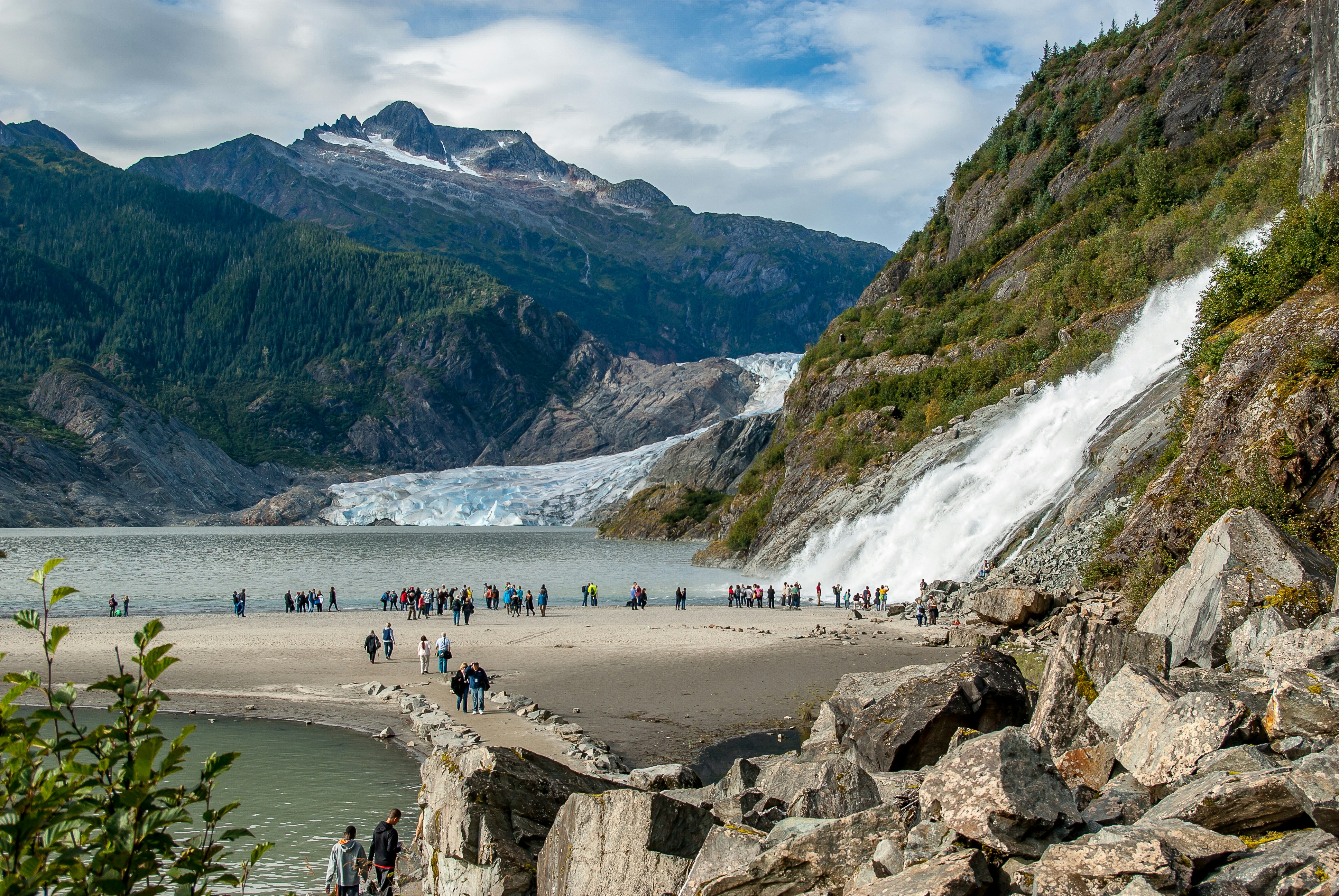 Visitors explore a sandy beach by a cascading waterfall and glacier under a vibrant blue sky.