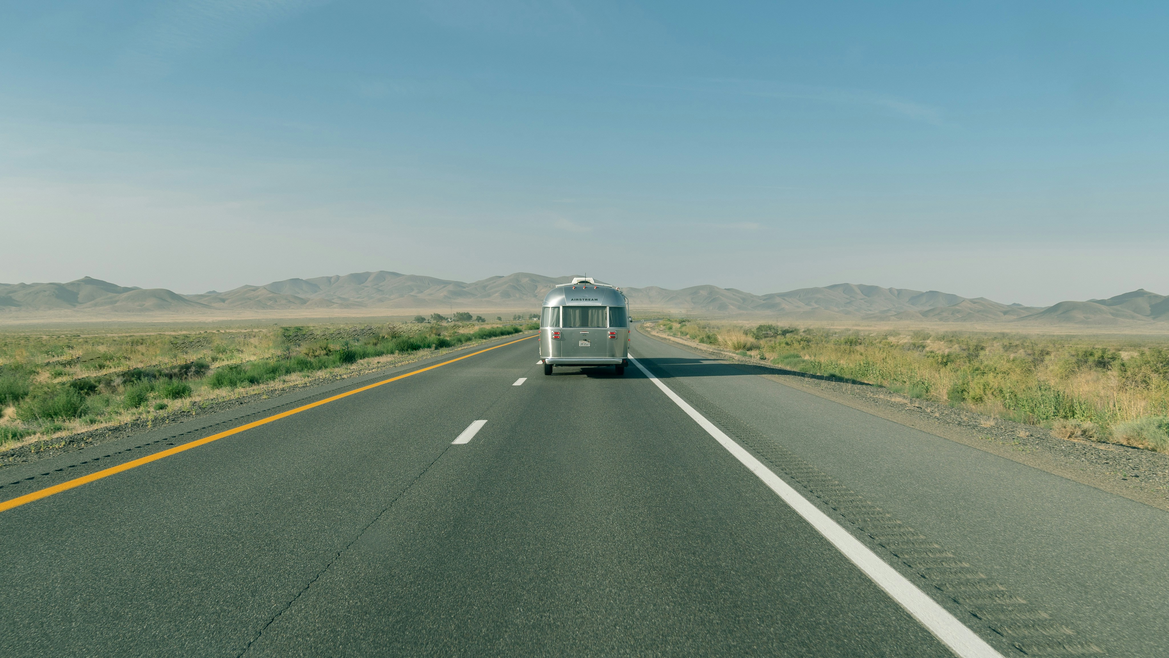 A silver Airstream trailer travels along an open highway, flanked by expansive grasslands and distant mountains under a clear blue sky.