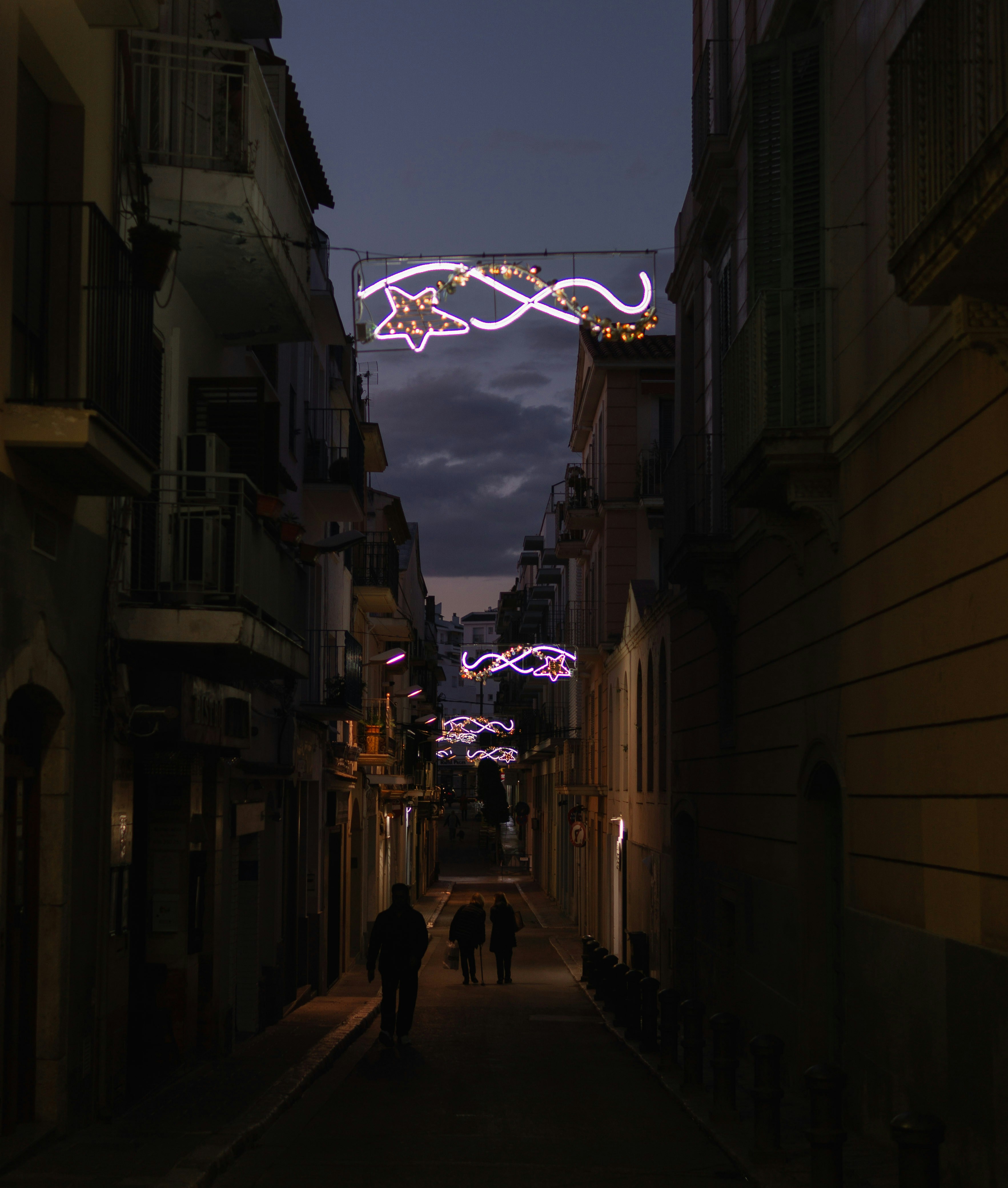 Couple walking down a narrow street adorned with vibrant holiday lights against a dusky sky.
