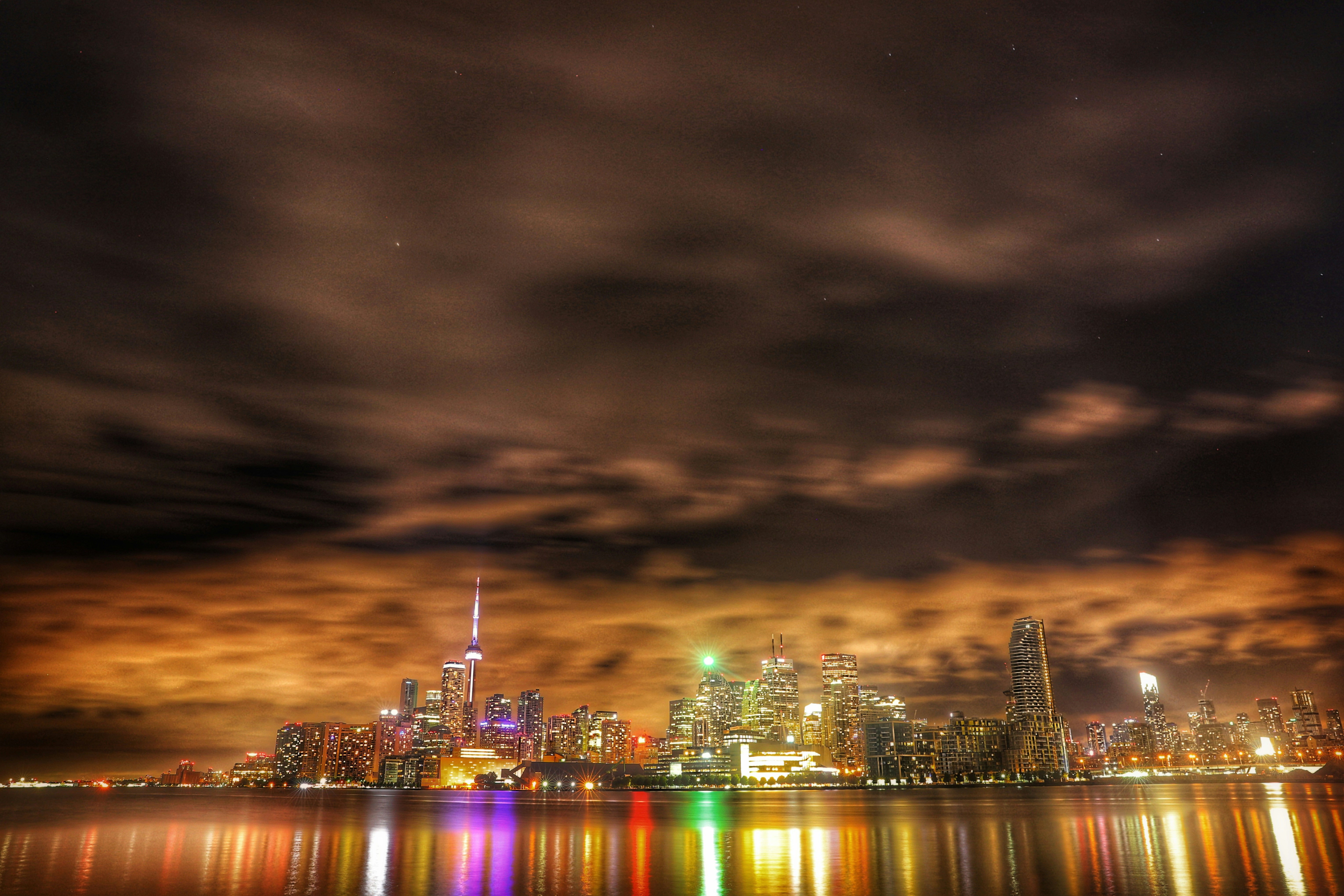 Vibrant city skyline illuminated by colorful lights reflecting on calm waters under a dramatic sky.