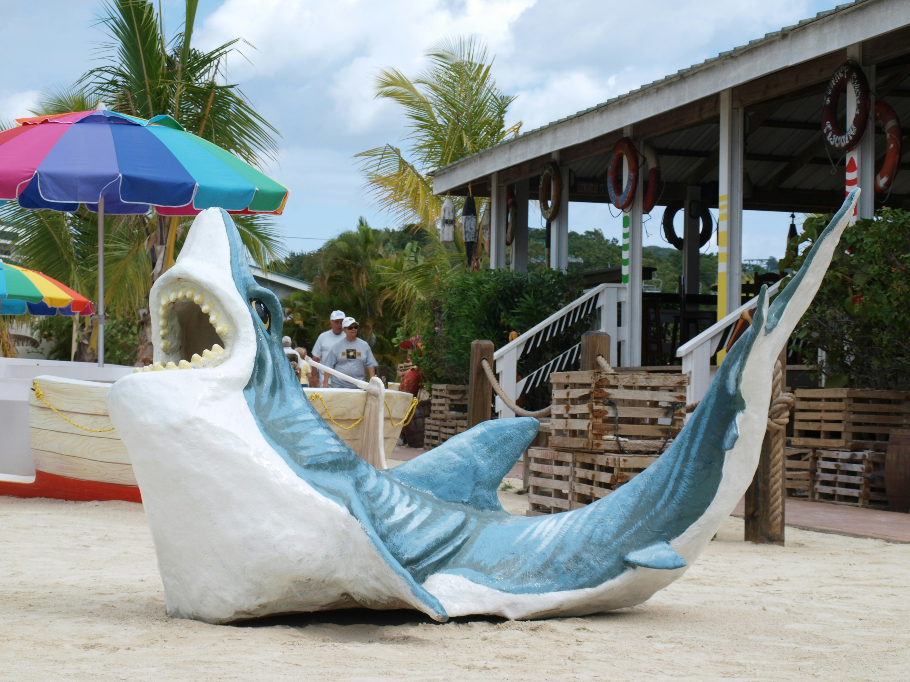 A large sculpted shark head sits on the sandy beach near a wooden boardwalk, with palm trees and colorful umbrellas in the background.