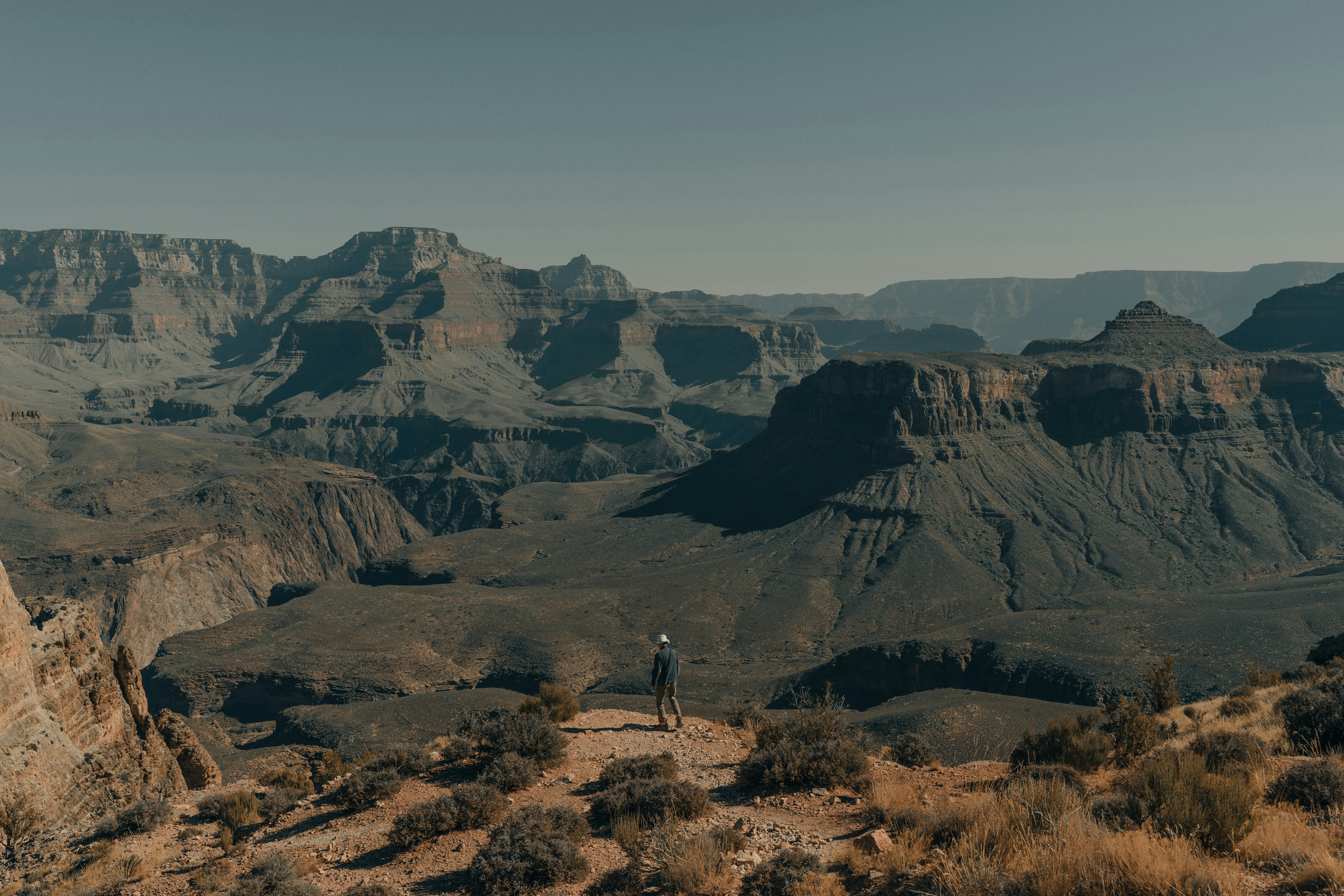 Hiker traverses a vast, arid canyon under a clear sky.