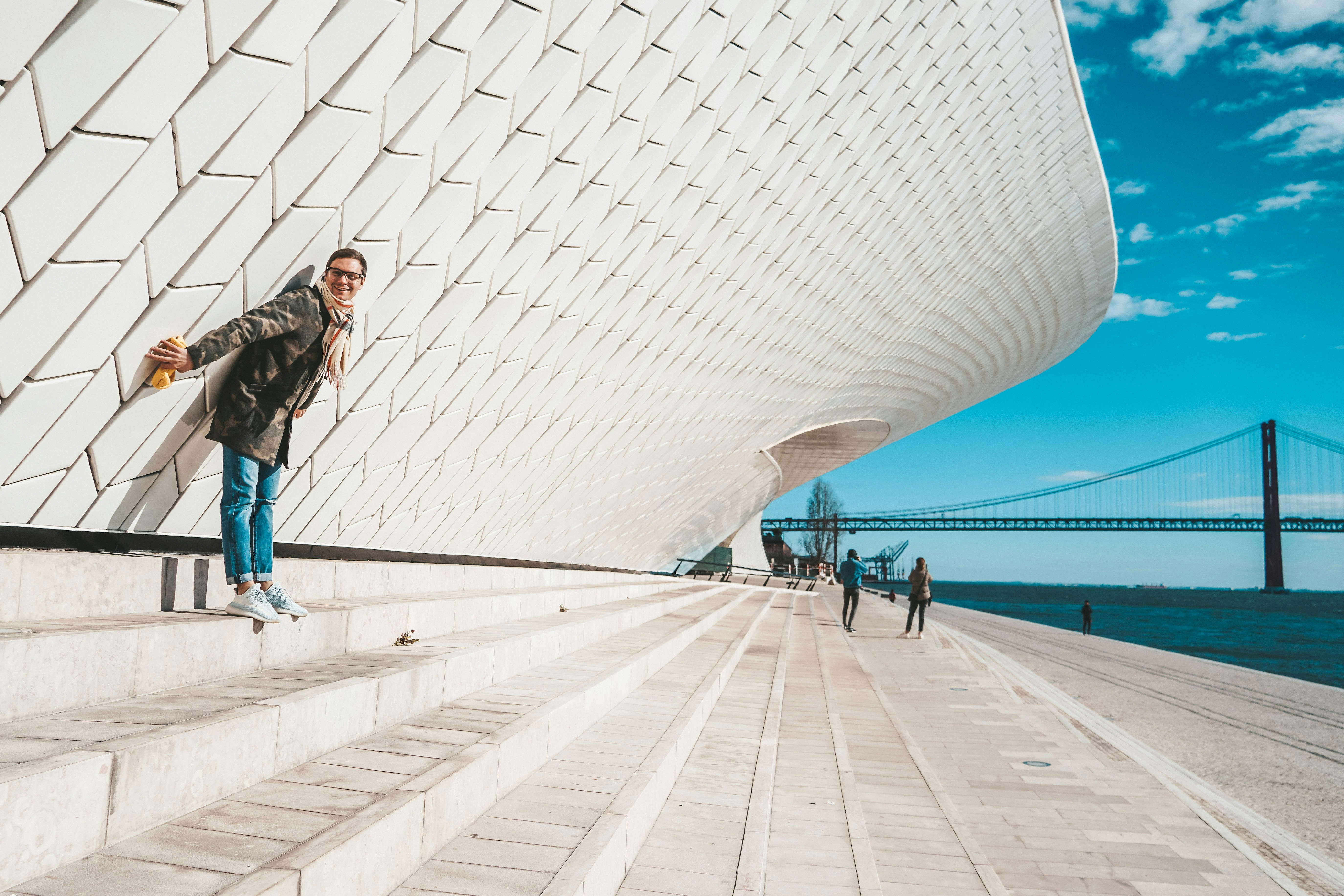 A person playfully leans against a modern architectural structure, with a bridge and waterfront in the background. The scene captures a blend of urban design and natural beauty.