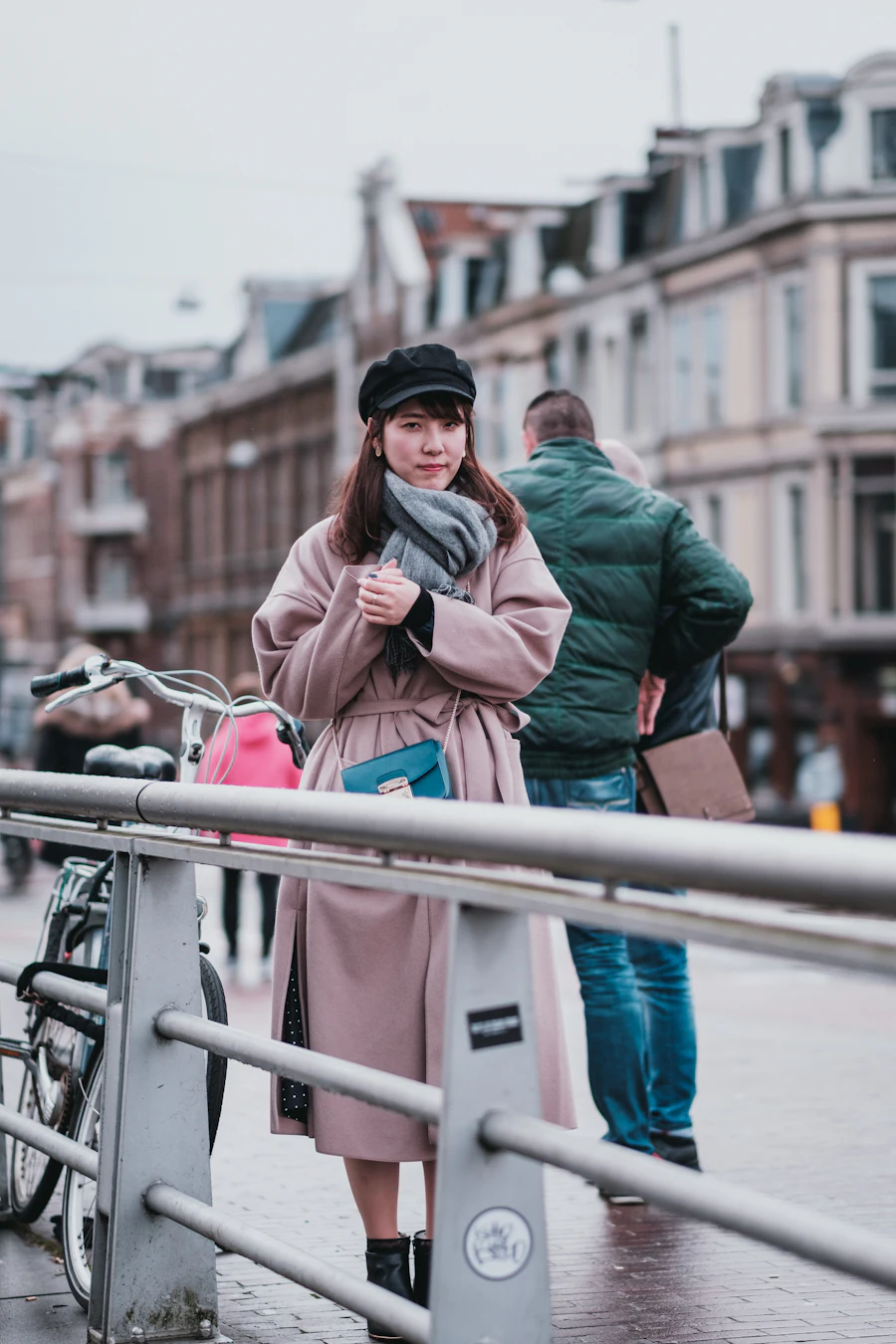 Young woman in a black cap and gray scarf stands by a railing on a rainy city street, with bicycles and blurred buildings behind her. A man in a green jacket stands in the background.
