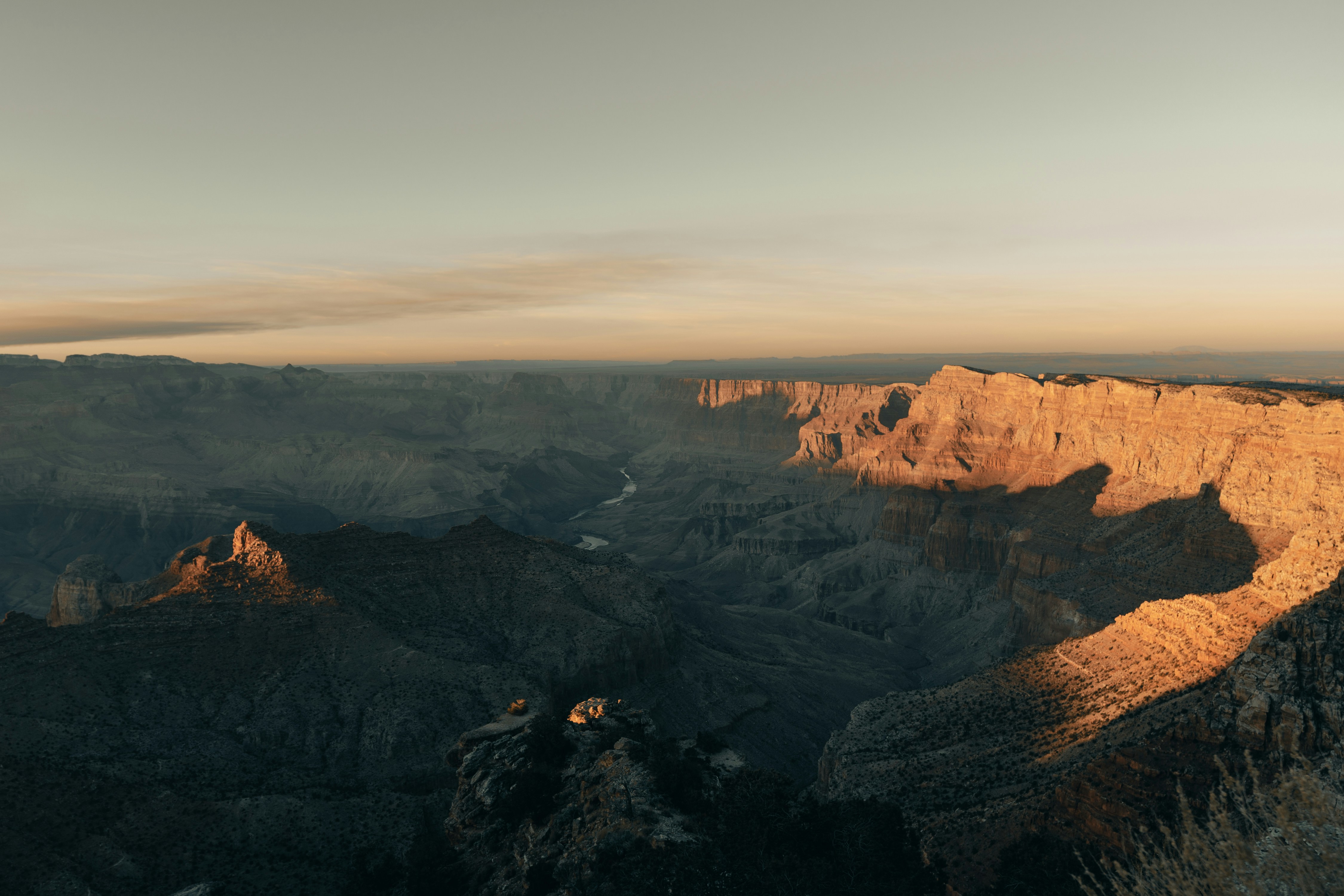 Golden light casts shadows over the rugged terrain of a canyon, revealing the intricate layers of rock formations. A river meanders through the depths, hinting at the landscape's vastness.