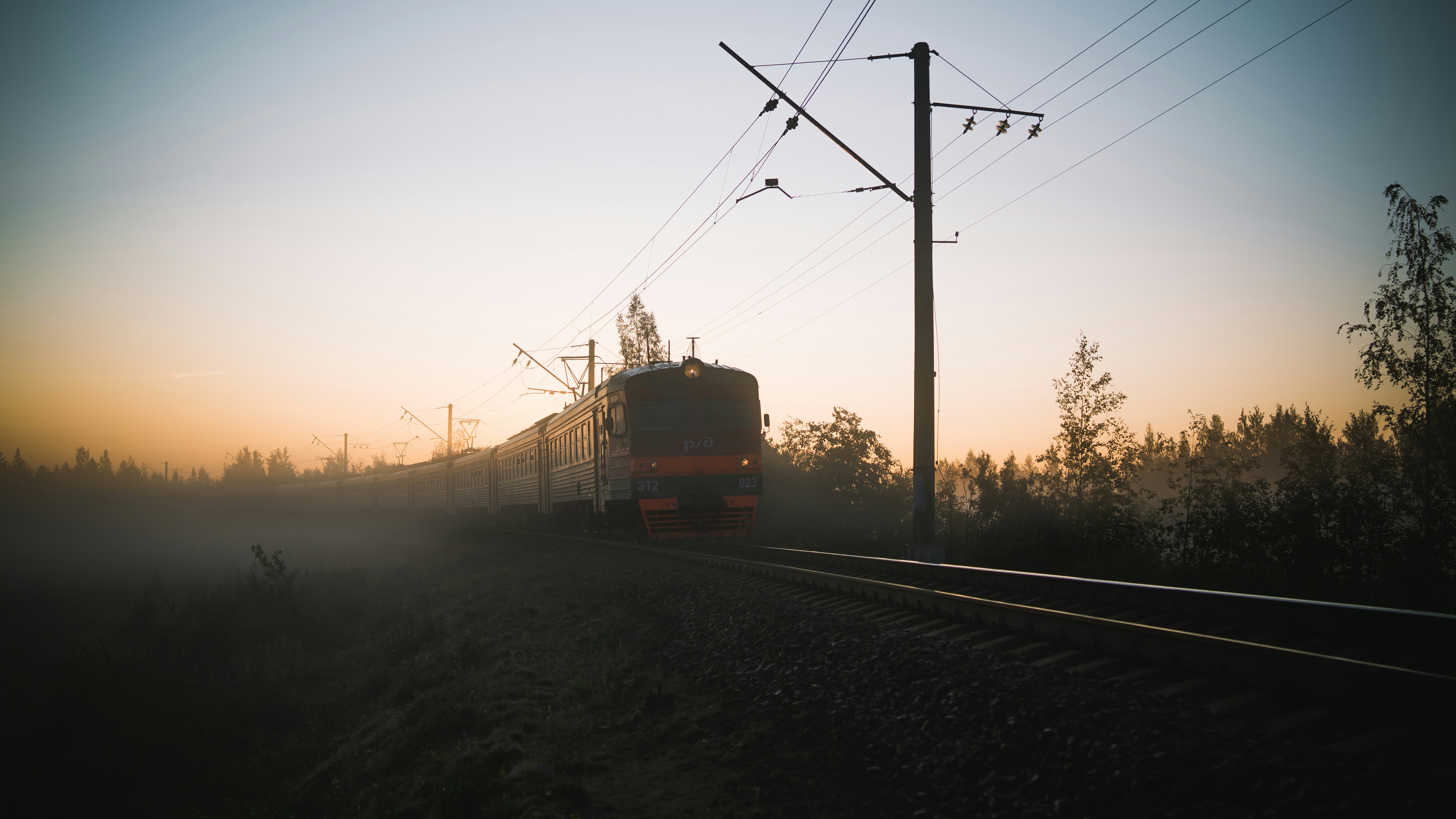 A train emerges from the fog at dawn, its silhouette contrasting against the soft pastel sky. Power lines stretch above, framing the scene.
