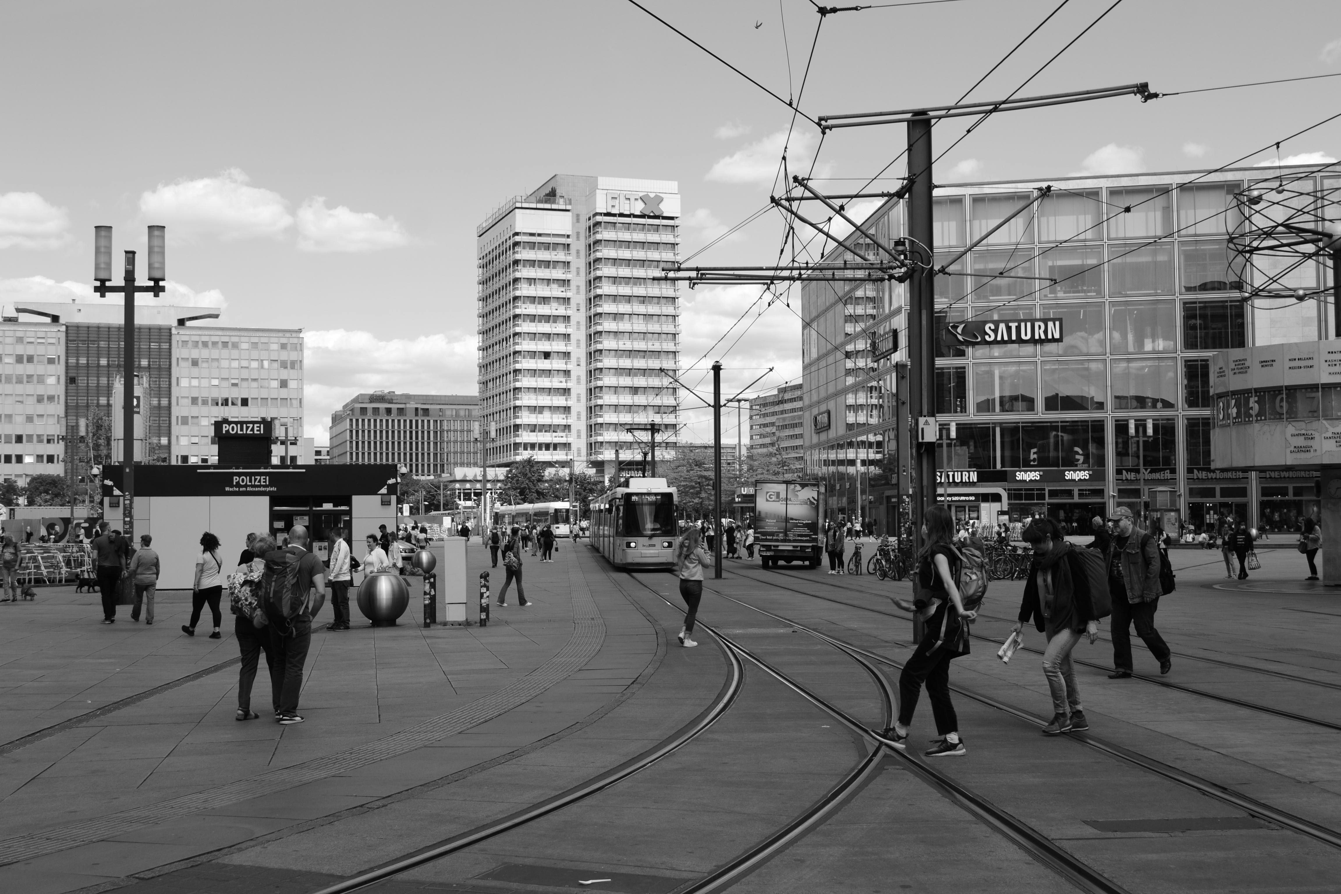 A bustling city intersection with pedestrians navigating tram tracks, surrounded by modern architecture and urban elements.